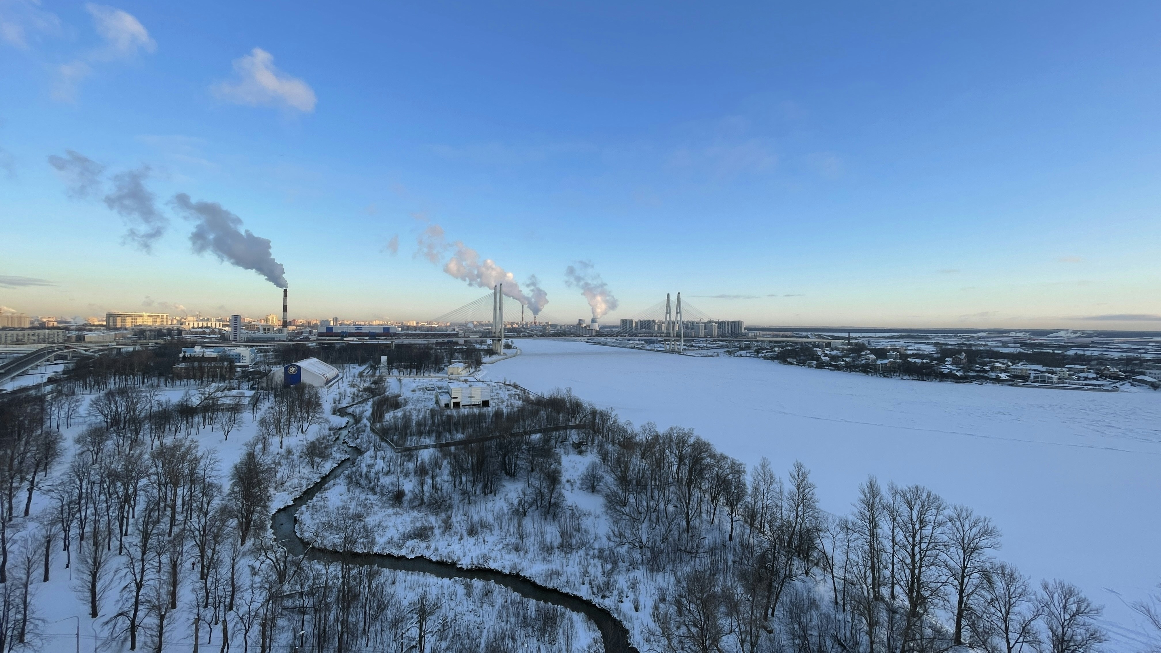 Industrial landscape with smoke billowing from chimneys against a clear blue sky, surrounded by snow-covered terrain and a frozen river.