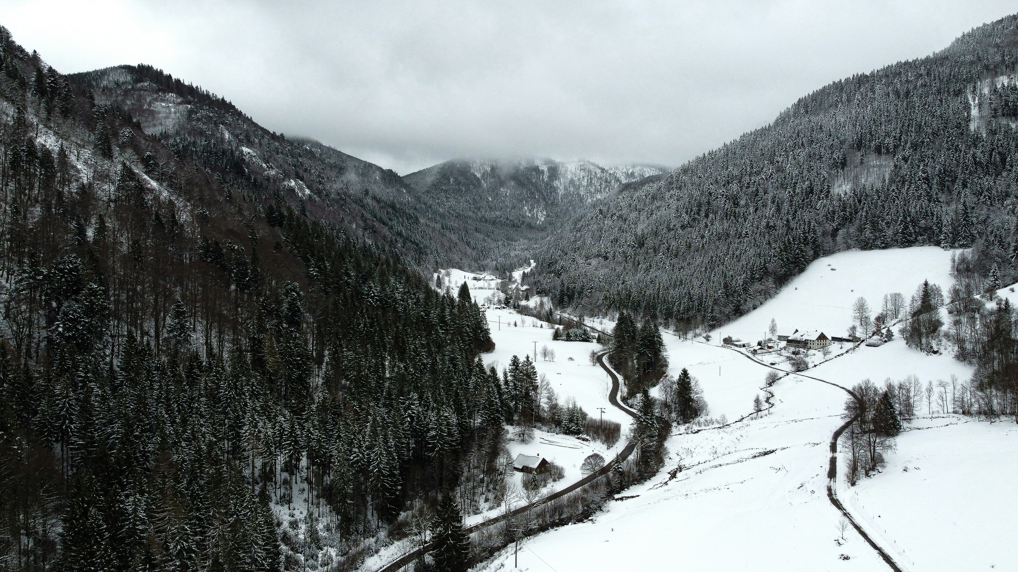 Snow-covered valley with winding river and scattered houses, framed by towering mountains under a cloudy sky.