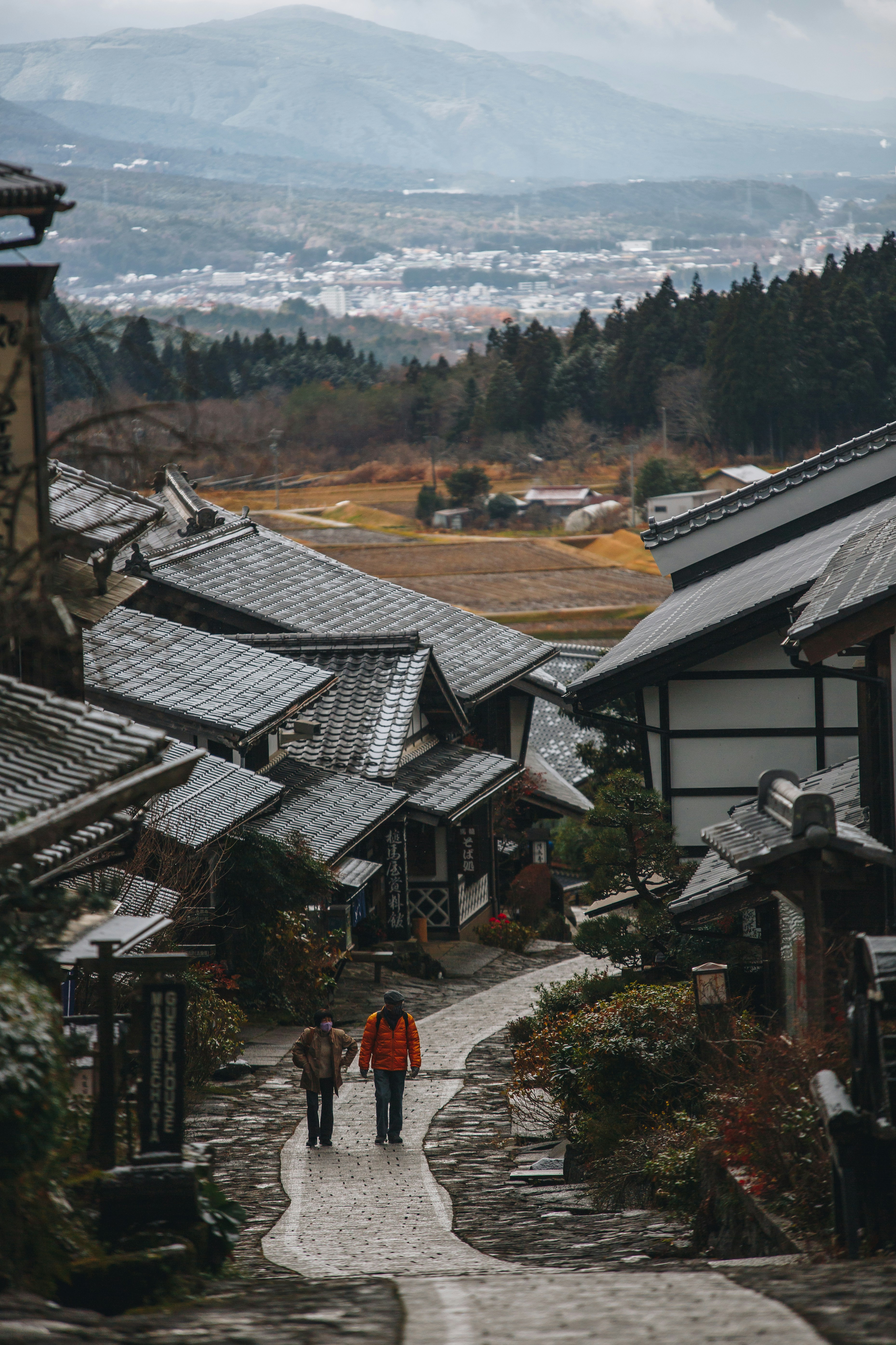 Couple walking down a cobblestone path flanked by traditional Japanese houses, with a backdrop of mountains and fields.