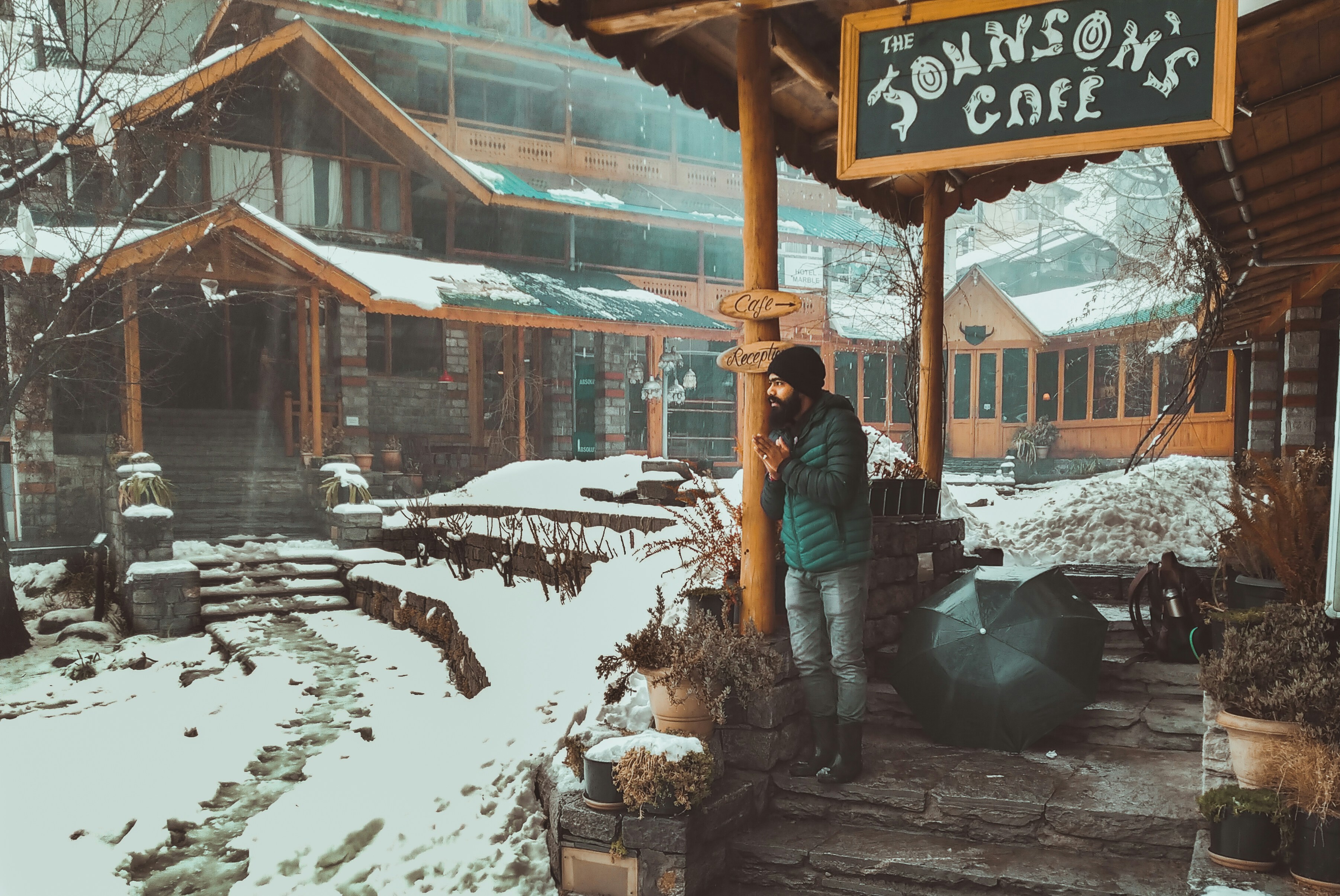 Cozy café nestled in a snowy landscape, featuring a visitor enjoying the ambiance under a rustic sign. Snow blankets the ground, enhancing the serene atmosphere.