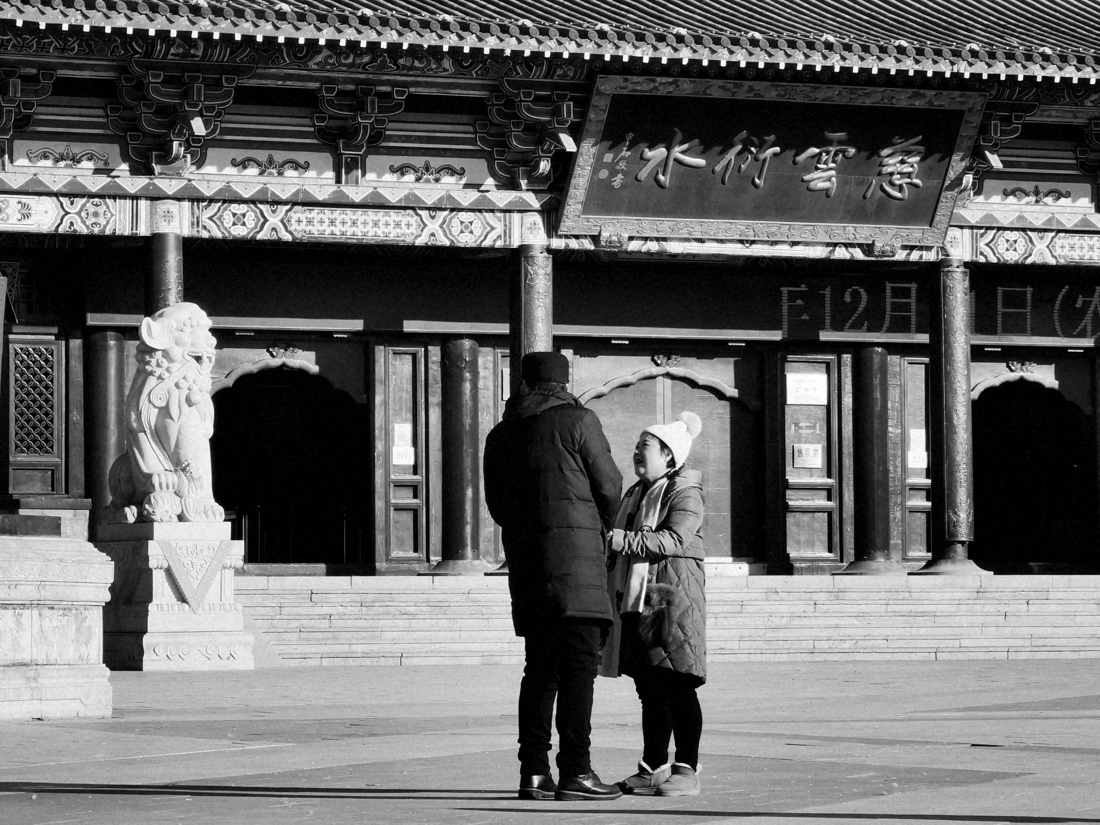 Two people stand in conversation before an ornate temple entrance in black and white.