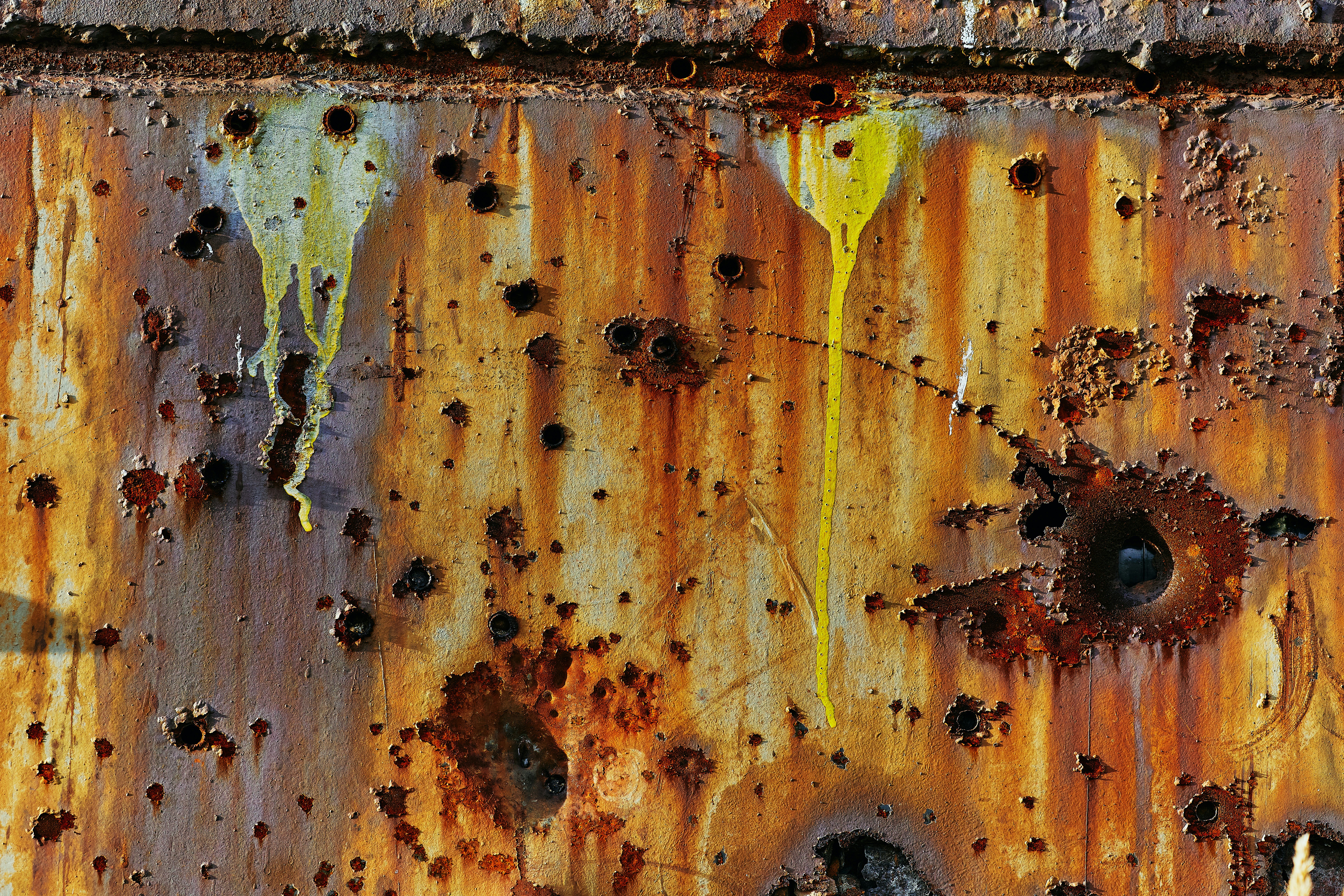 A rusty metal wall with splashes of yellow and orange paint in close-up. Metal wall with rust and bullet holes. Textured metal background with cracks, bullet holes and yellow paint