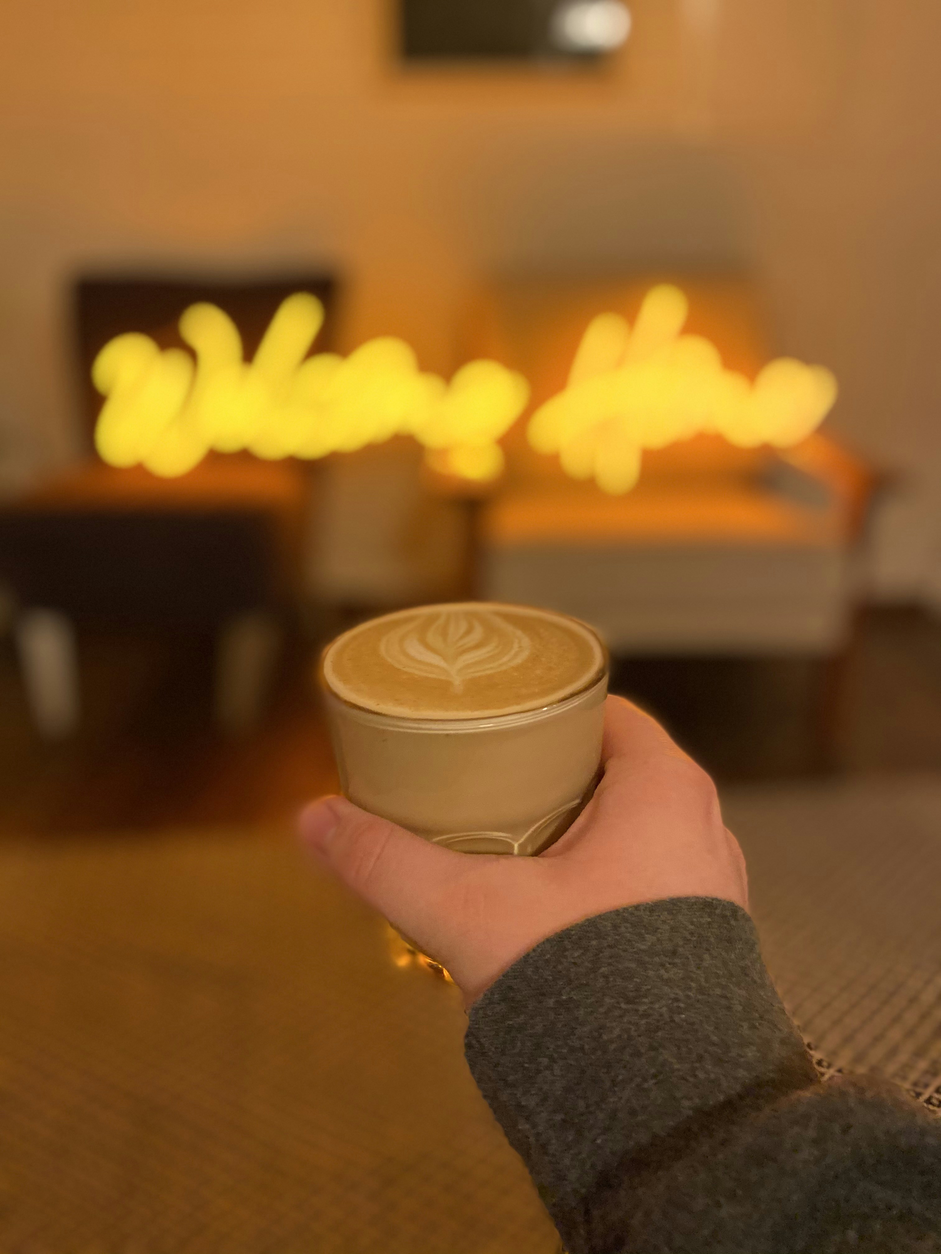 Latte art displayed in a hand-held cup, with soft glowing text in the background creating a warm atmosphere.
