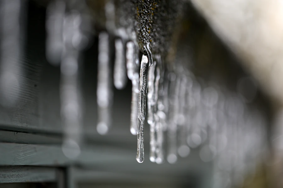 A row of icicles hangs from a dark roof edge, with one long icicle sharply in focus near the center. The background falls into soft blur with pale light and more ice shapes receding into the distance.