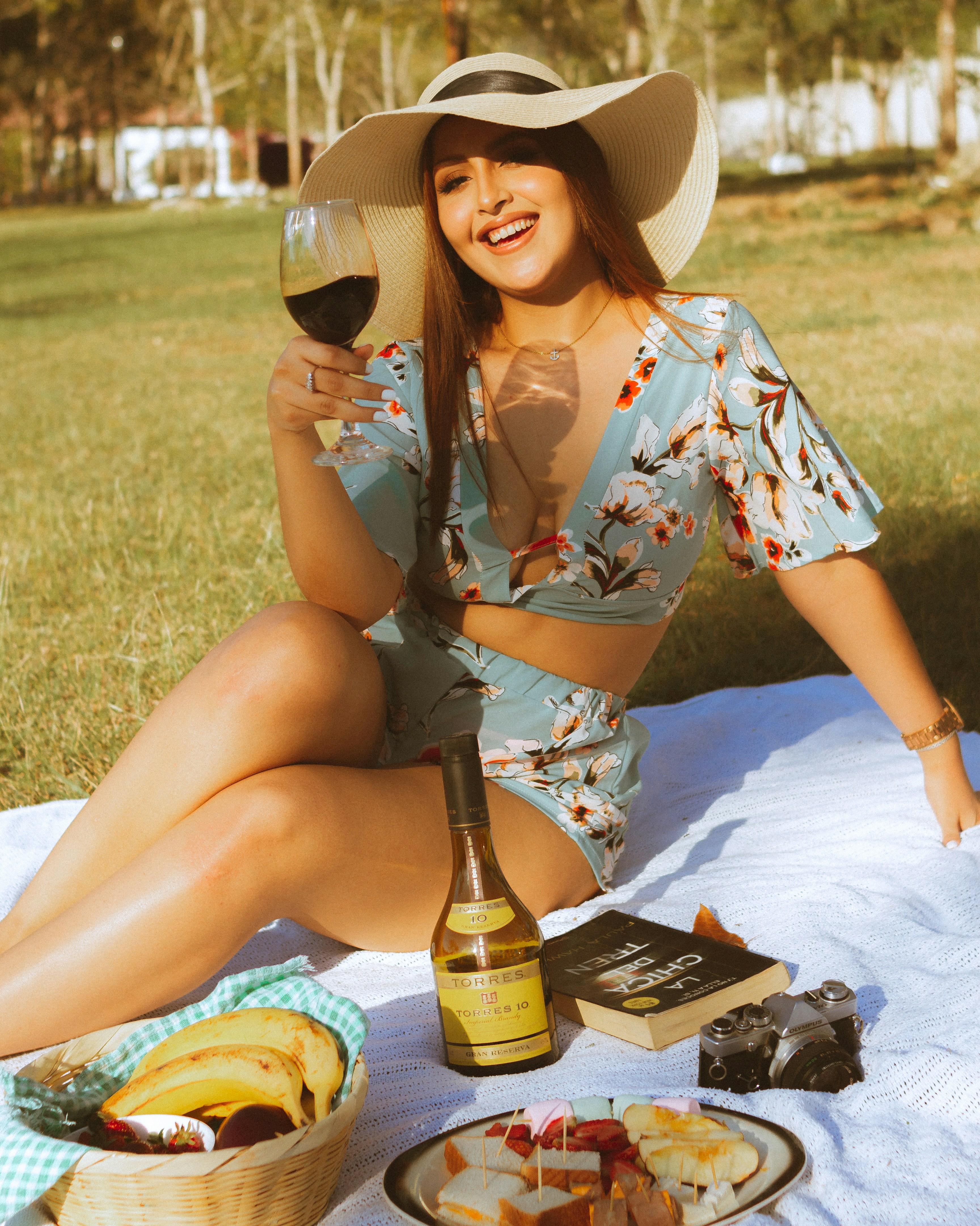 Woman in a floral blue two-piece dress and wide-brim hat sits on a blanket with wine, fruit, and camera on a sunny lawn.
