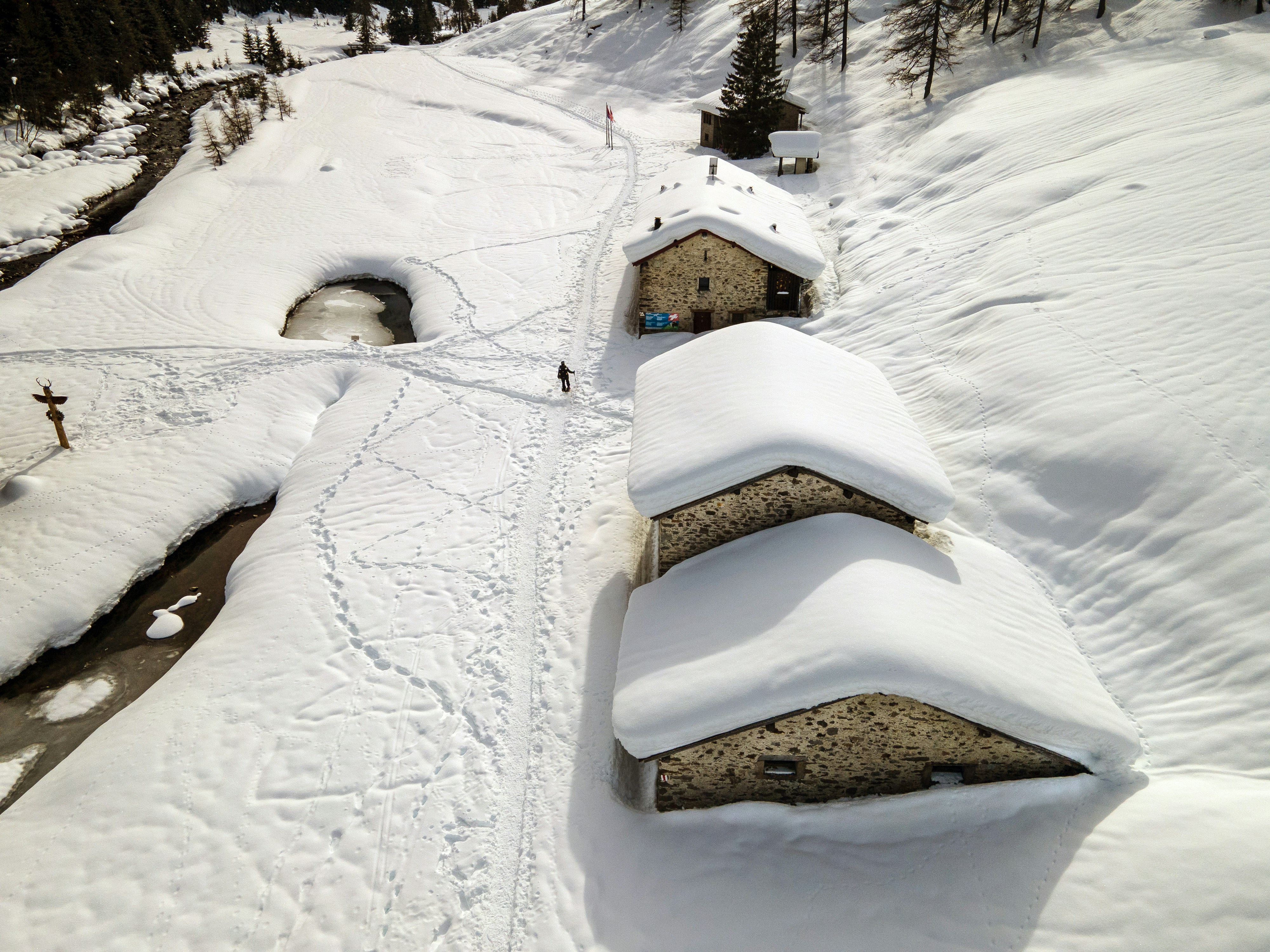 Two stone cabins blanketed in snow beside a winding path and a small pond, evoking a serene winter landscape.