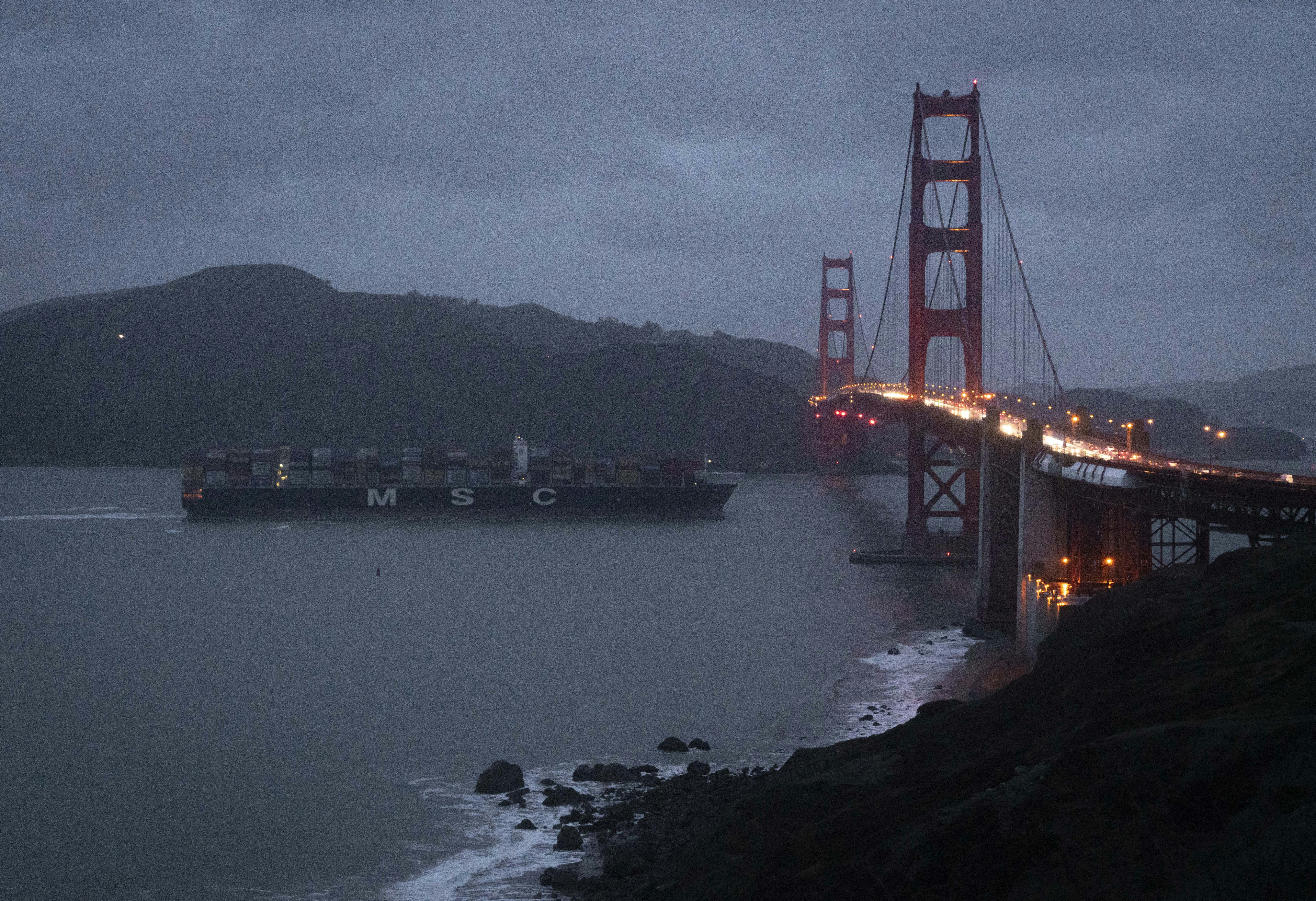 Container ship navigating under the Golden Gate Bridge, shrouded in a moody twilight ambiance. The scene captures the interplay of light and shadow along the coastal landscape.