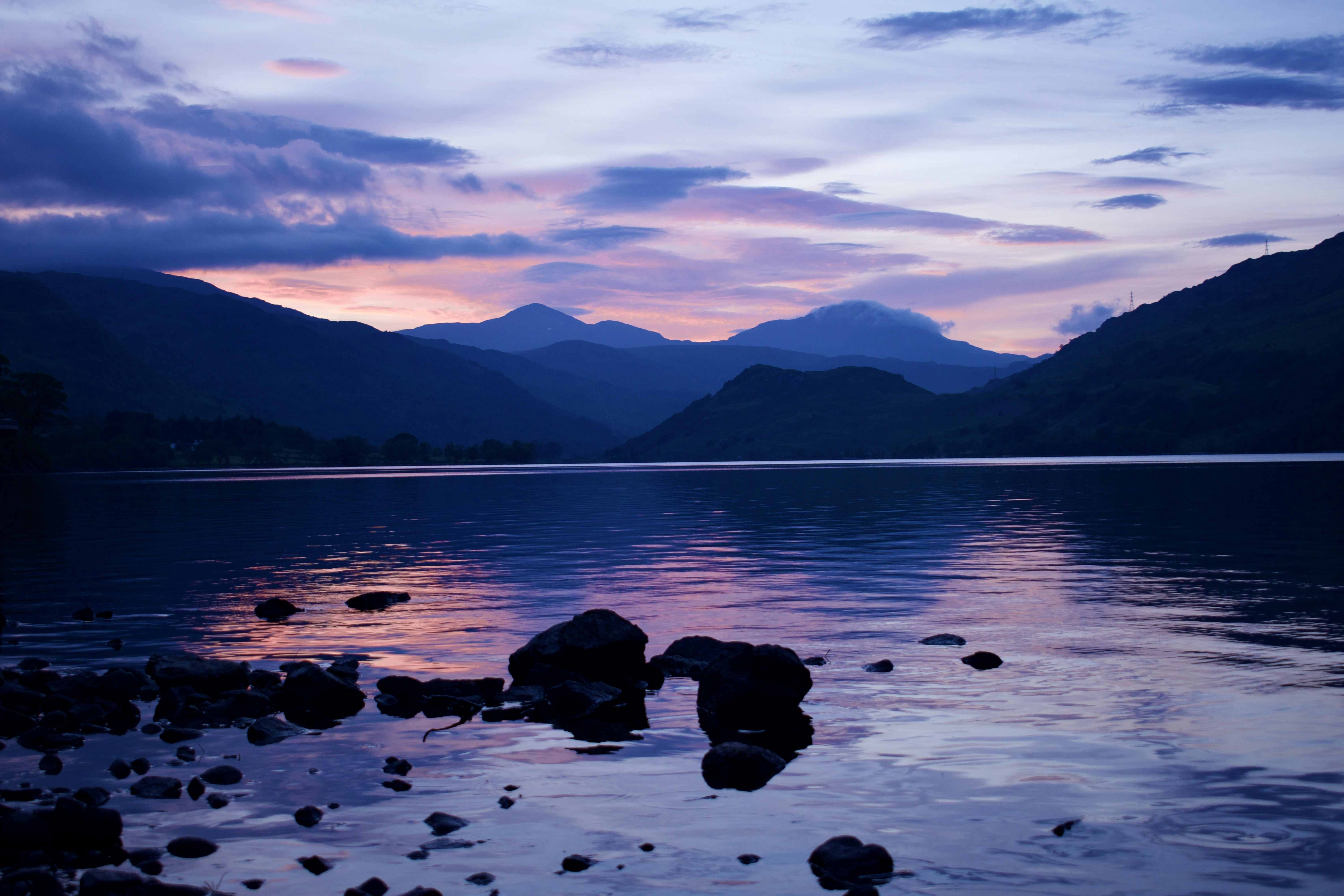 Calm lake reflecting twilight hues, framed by distant mountains and rocky shoreline.