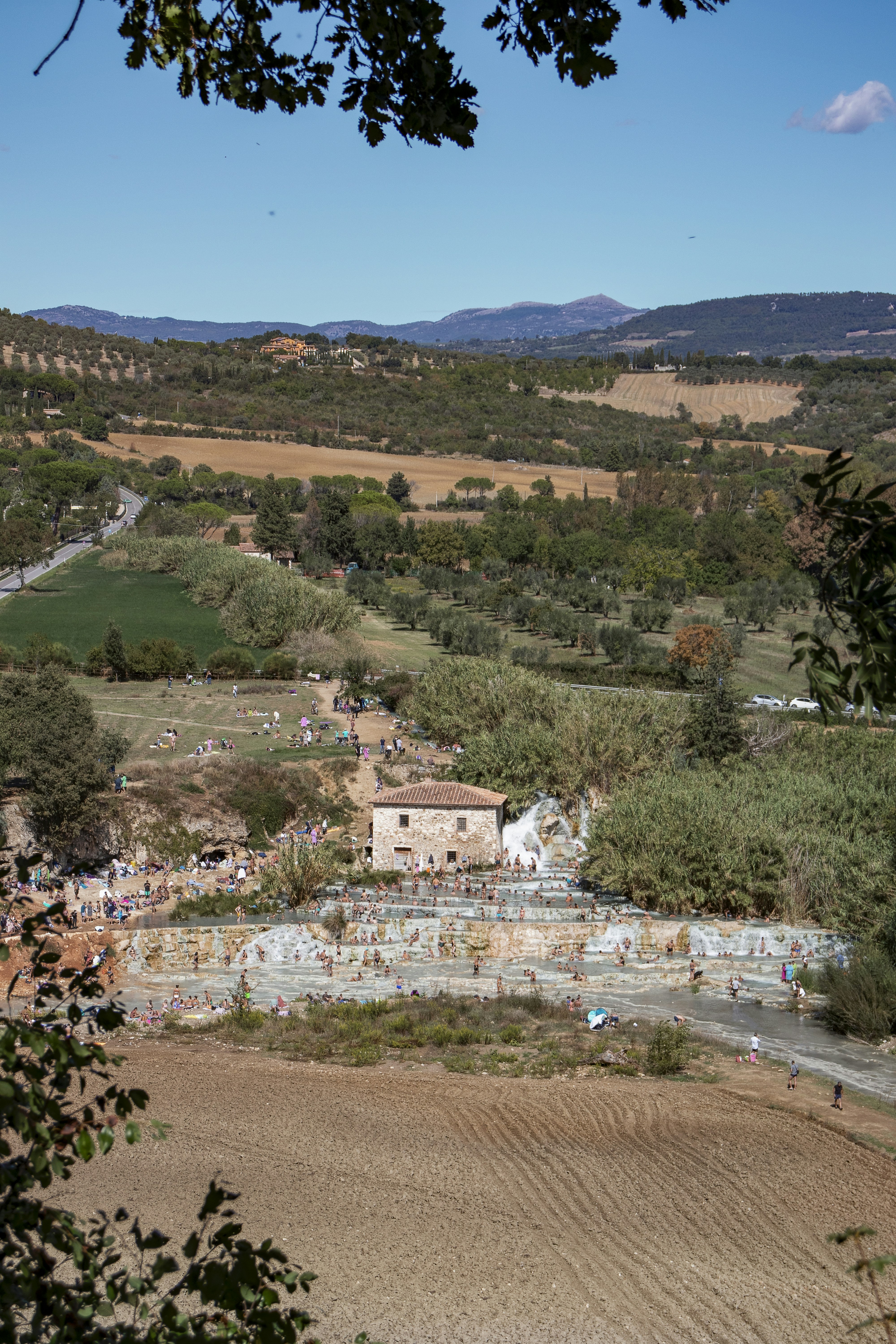 Visitors enjoying natural thermal springs surrounded by lush greenery and rolling hills. The quaint building adds charm to the tranquil landscape.