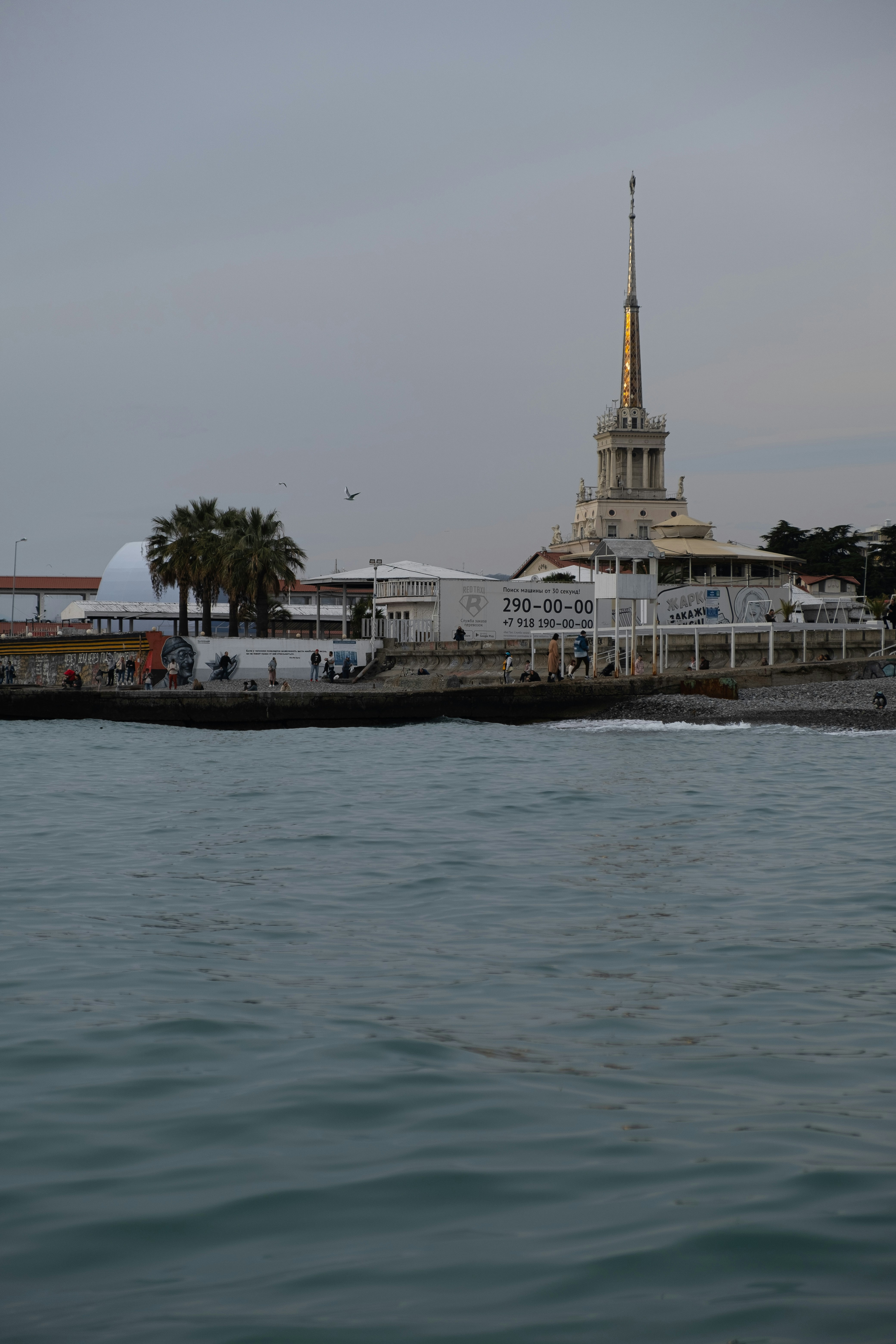 Historic coastal building with a tall spire overlooking the sea, surrounded by palm trees and visitors near the shoreline.