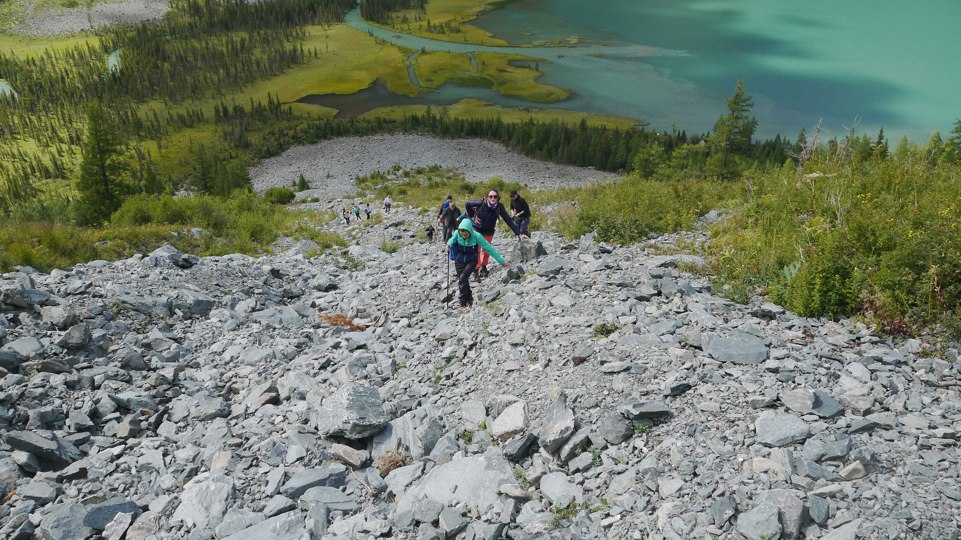 Hikers navigate a rocky slope with a vibrant turquoise lake and lush greenery in the background. The scene captures the challenge and beauty of outdoor exploration.