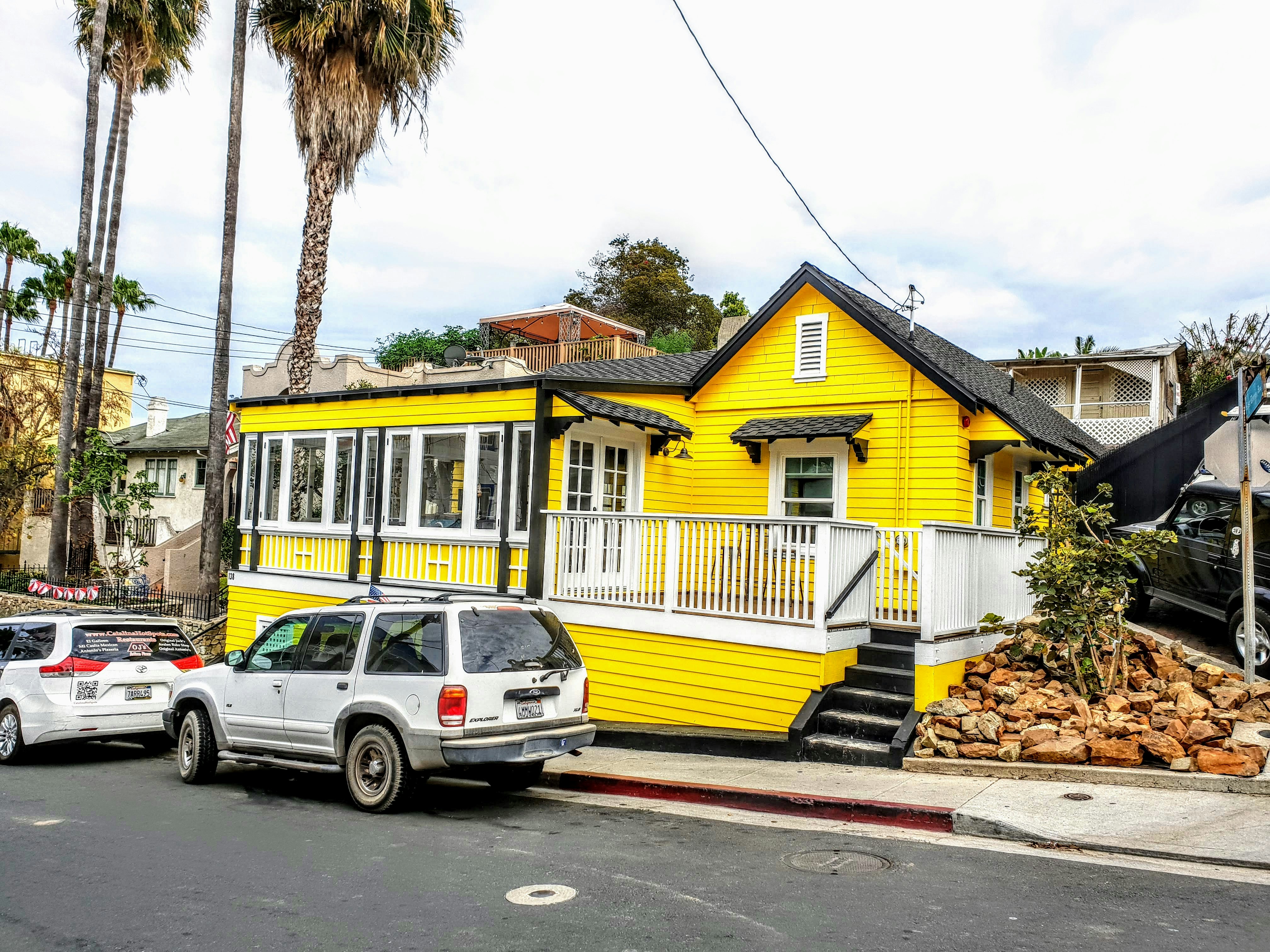 Bright yellow house with a modern design, surrounded by palm trees and parked cars on a sloped street.