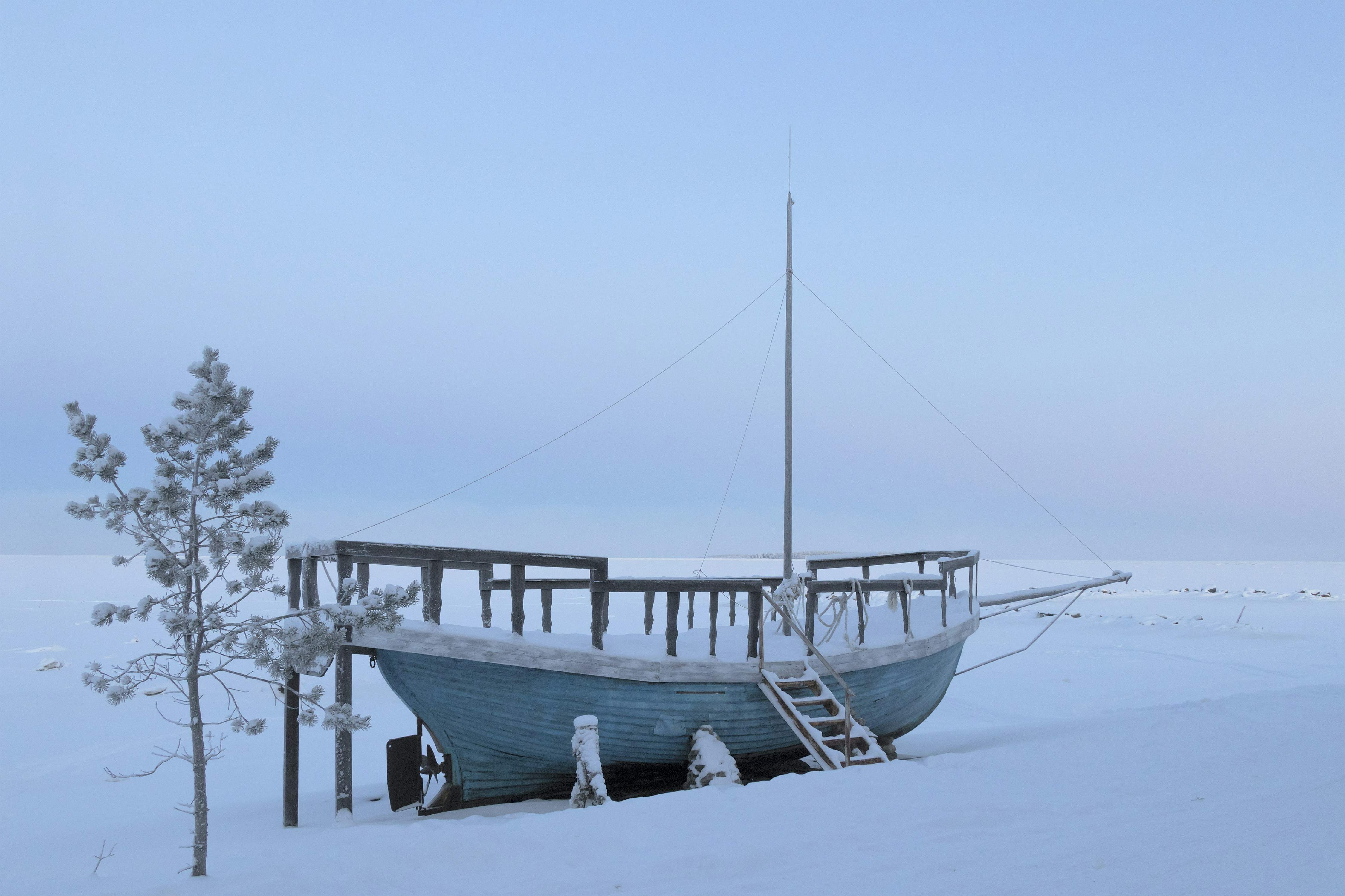 Abandoned wooden boat resting on a snowy landscape, surrounded by frost-covered trees under a pale sky.