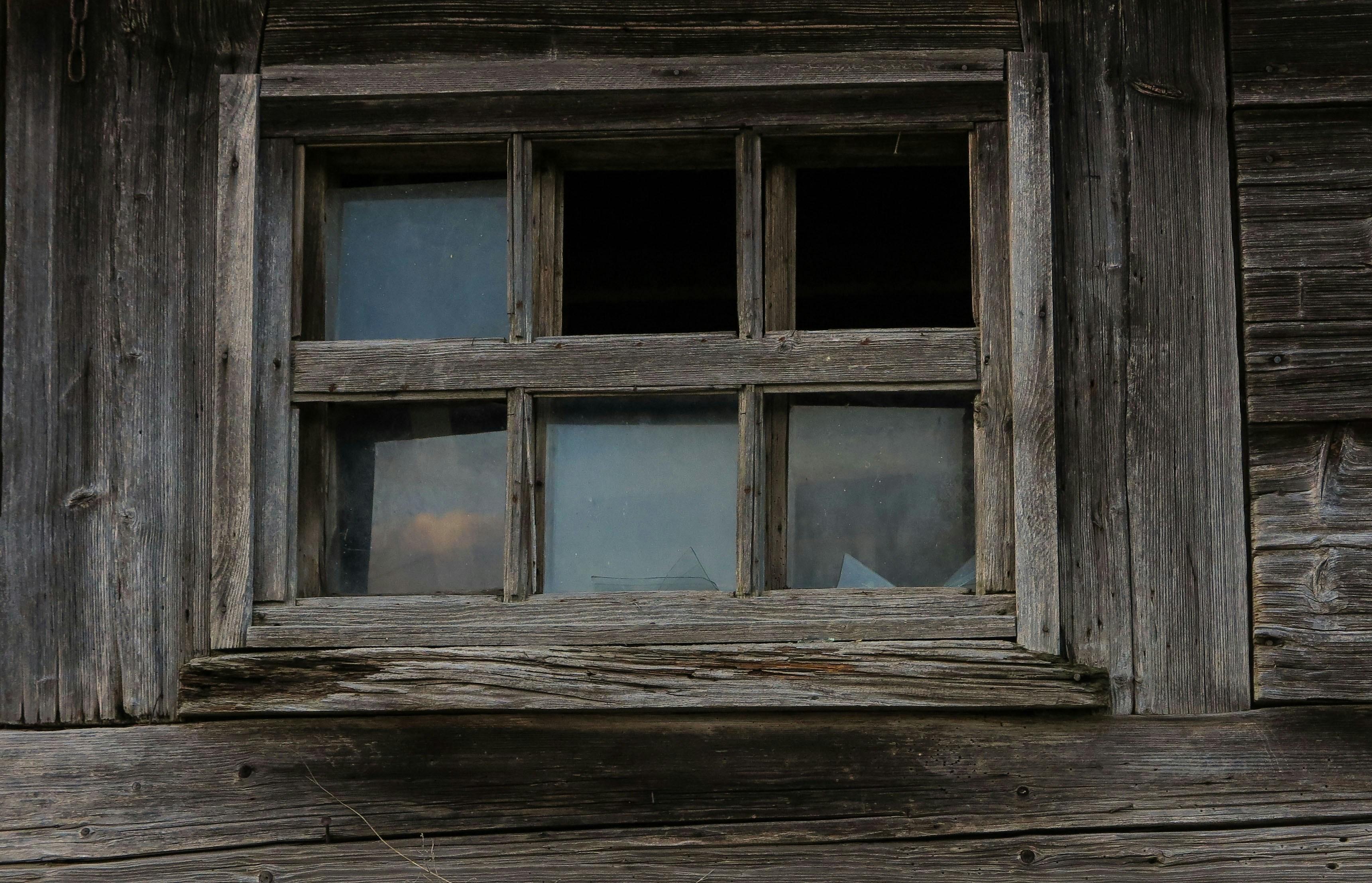 A weathered wooden window with nine panes sits in a rough timber wall, captured in muted, rustic light. The glass reflects a hint of the sky, suggesting age and quiet abandonment.