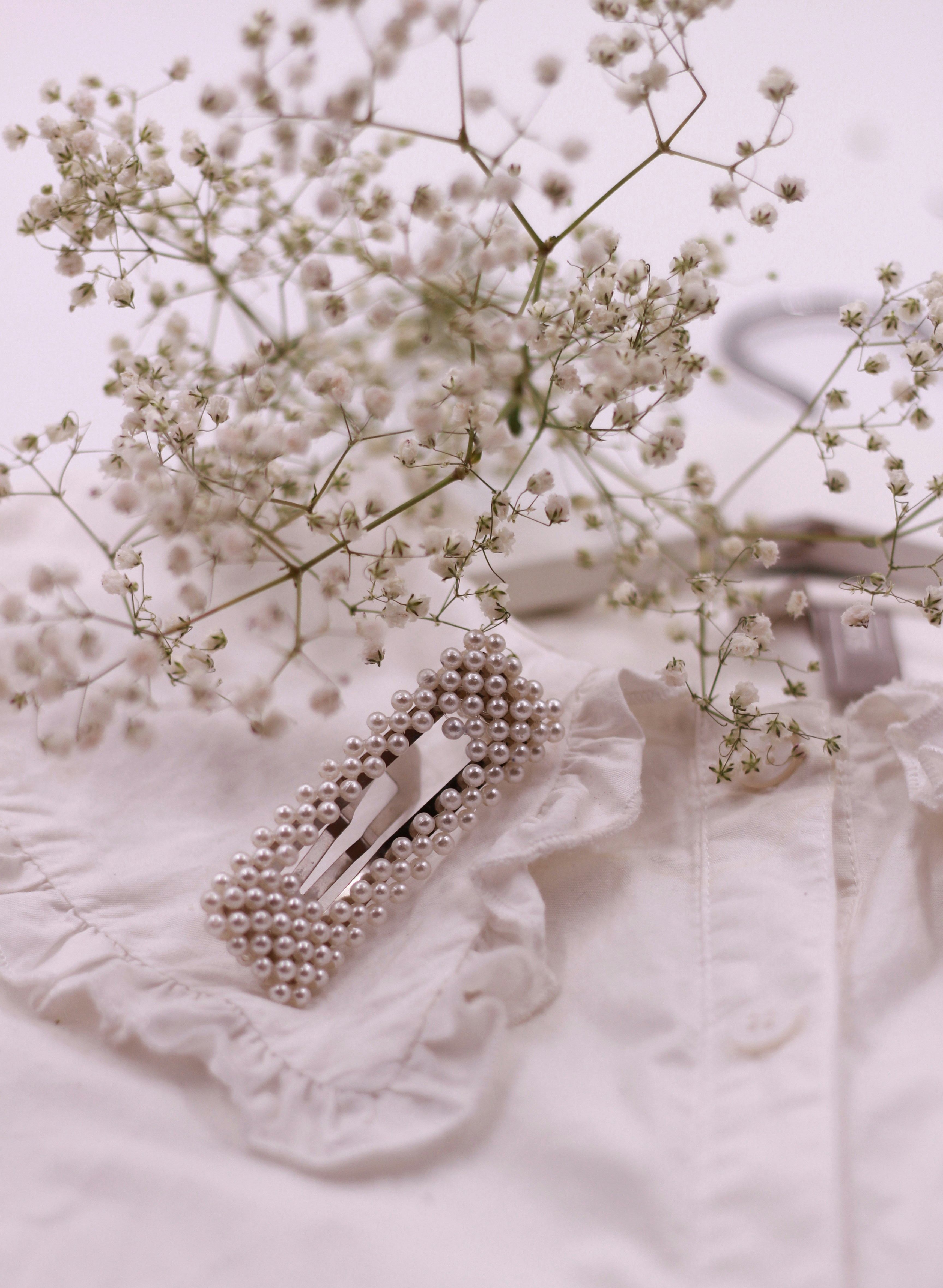 A pearl-embellished hair clip rests on a softly textured white blouse, surrounded by delicate gypsophila flowers, creating a serene and elegant ambiance.