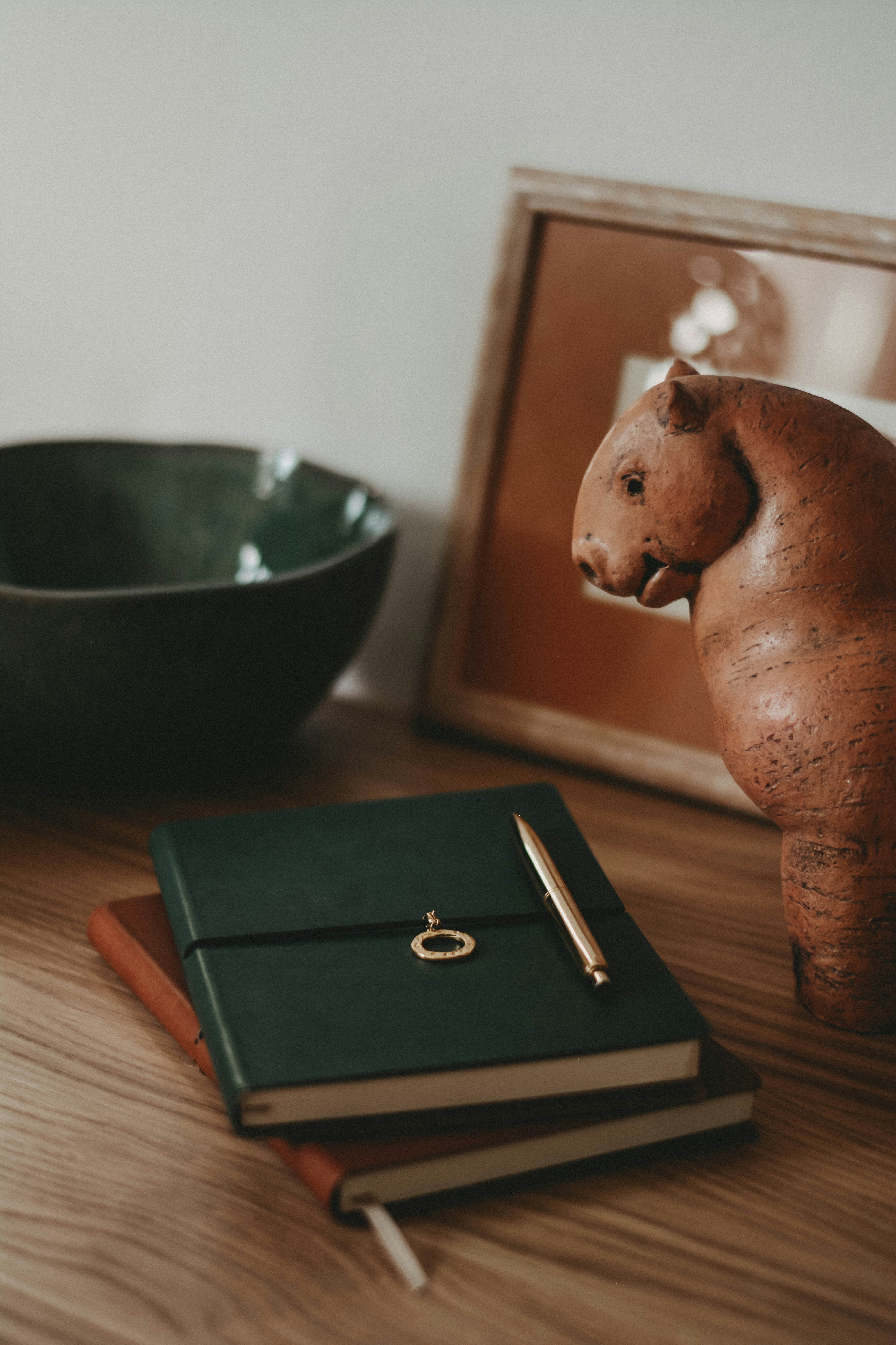 A green notebook rests atop a brown one, accompanied by a gold pen and a clay horse figurine, with a green bowl and a framed picture in the background.