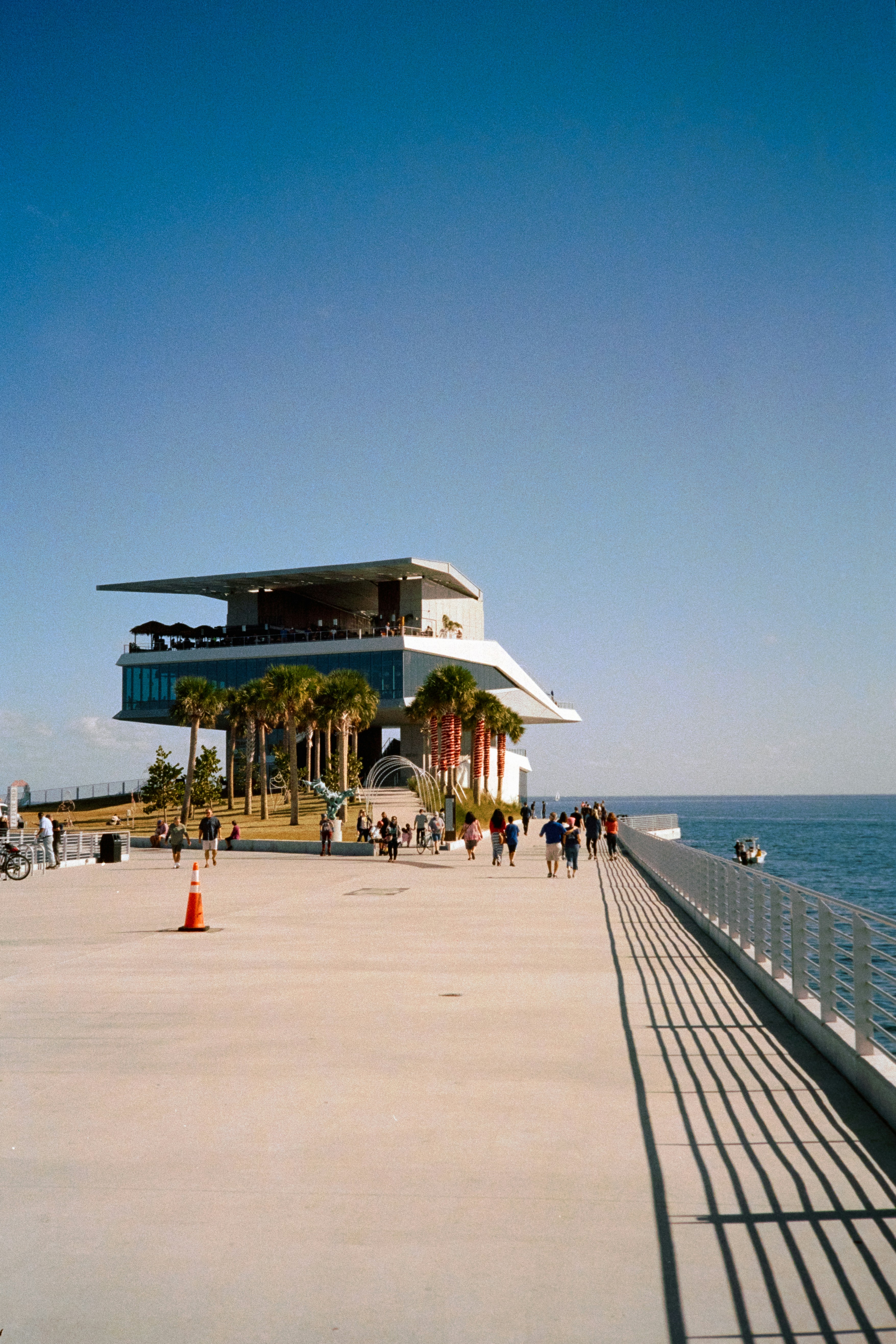 A contemporary building with a spacious terrace overlooks the ocean, surrounded by palm trees and visitors strolling along the waterfront promenade.