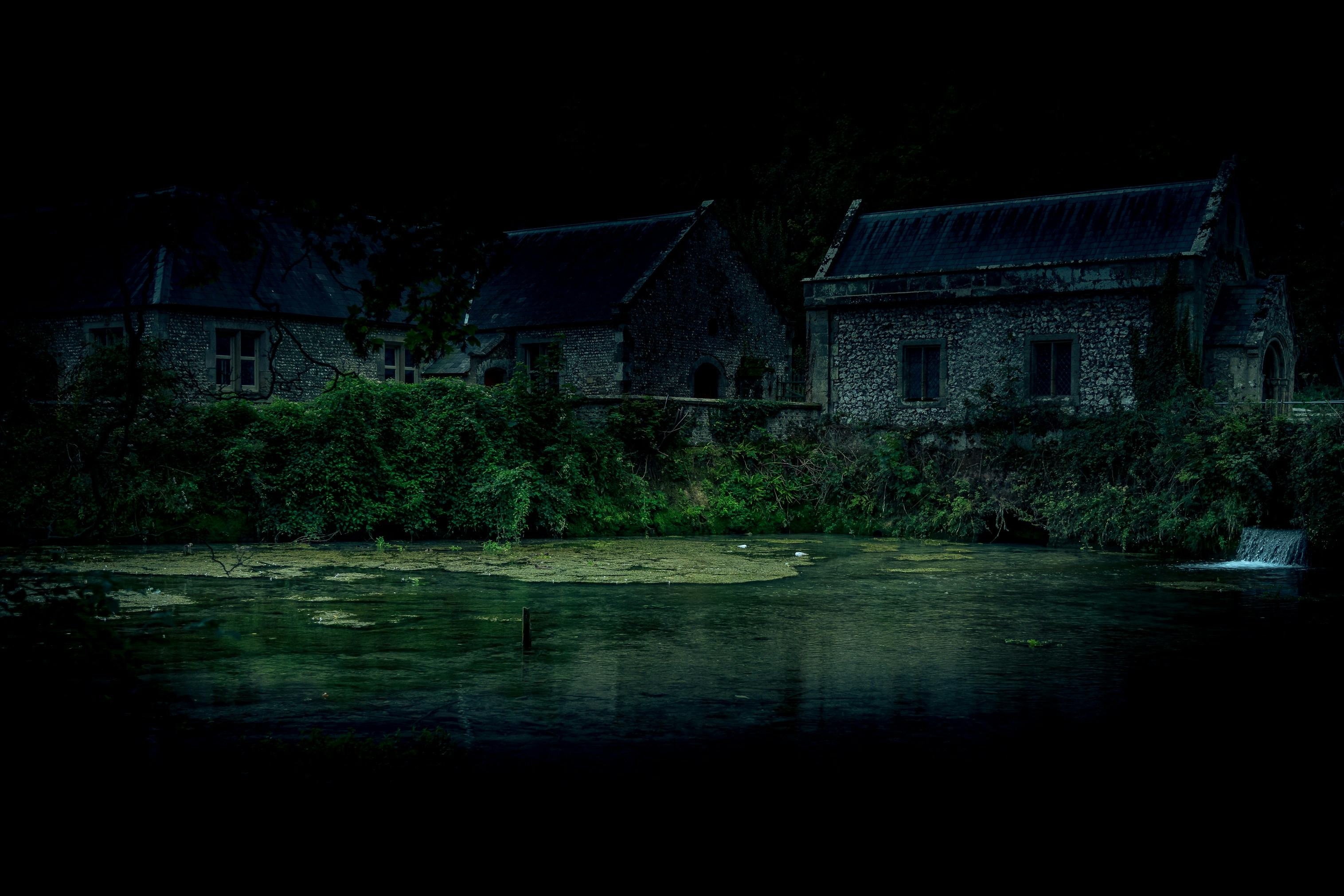 Dilapidated stone buildings shrouded in darkness beside a still, murky pond, hinting at a once-thriving location now lost to time.