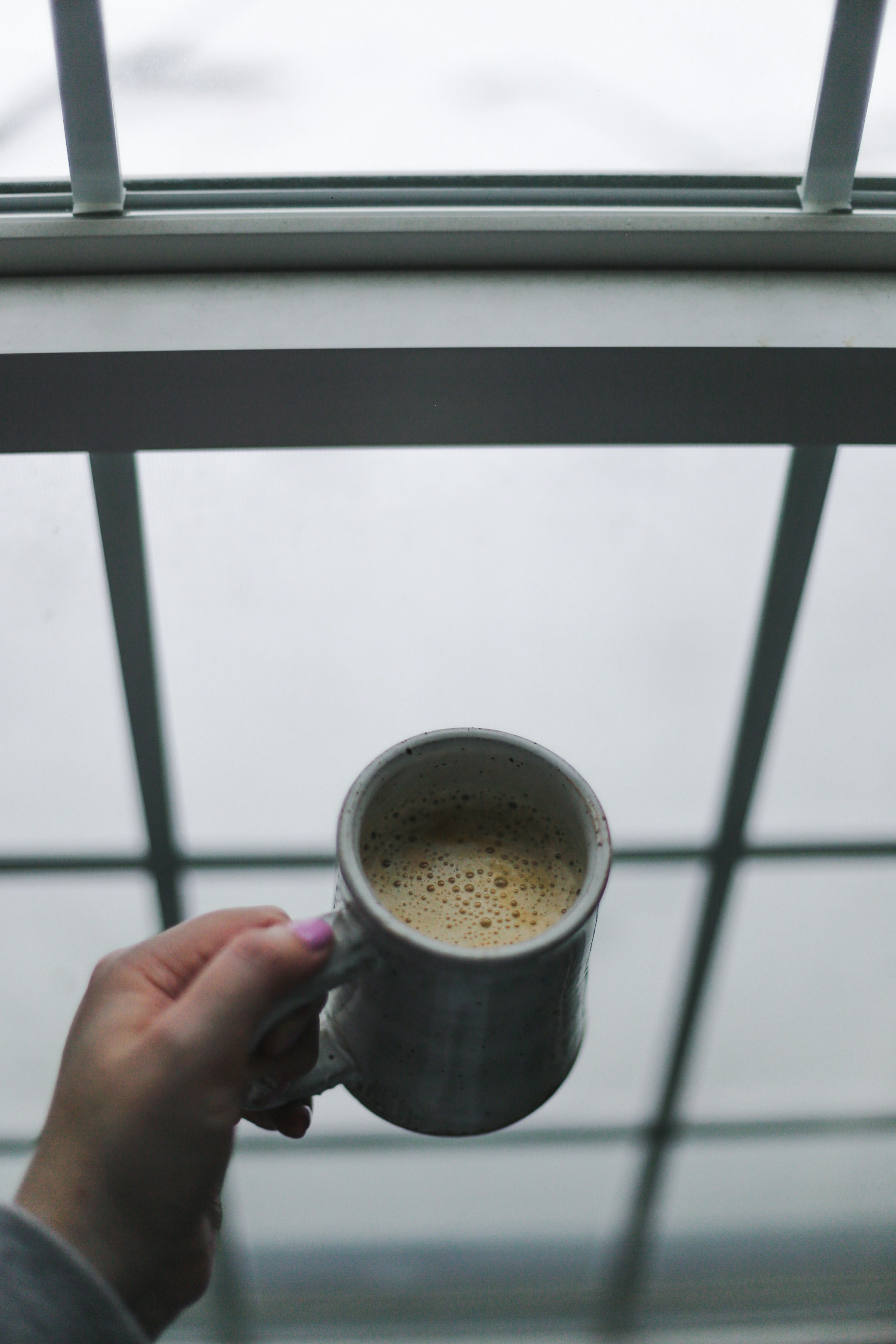 Hand holding a ceramic mug filled with frothy coffee, framed by a window with a soft, cloudy backdrop.
