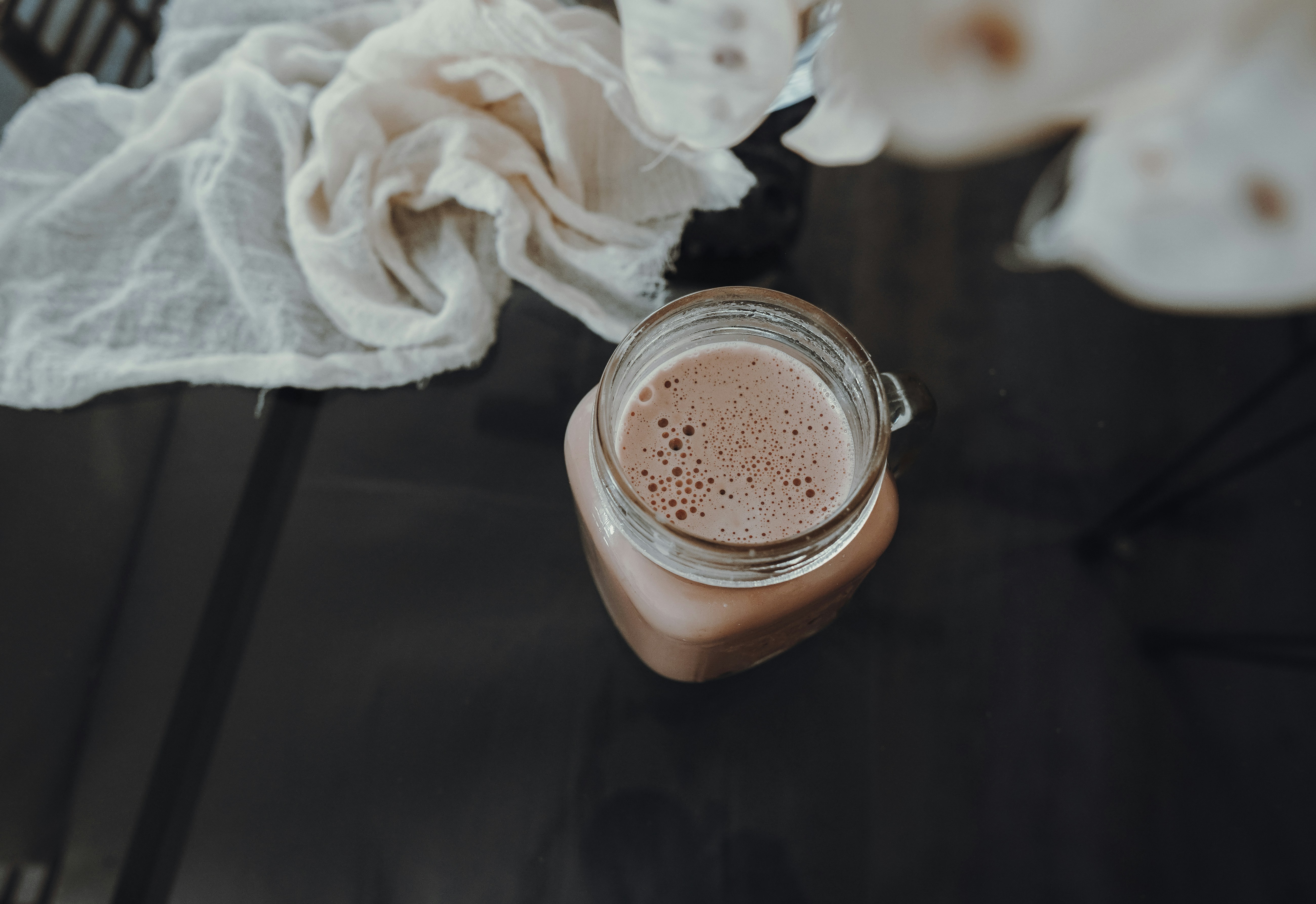 A glass jar filled with frothy beverage sits on a dark table, surrounded by soft fabrics and delicate floral elements.