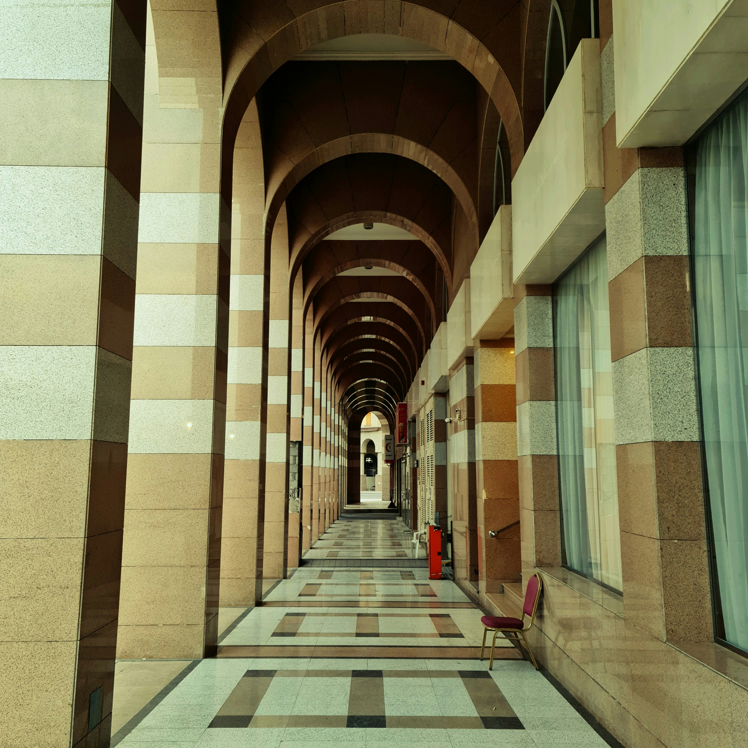 Elegant arched corridor lined with patterned tiles and soft lighting, leading to a distant doorway. A solitary chair adds a touch of human presence.