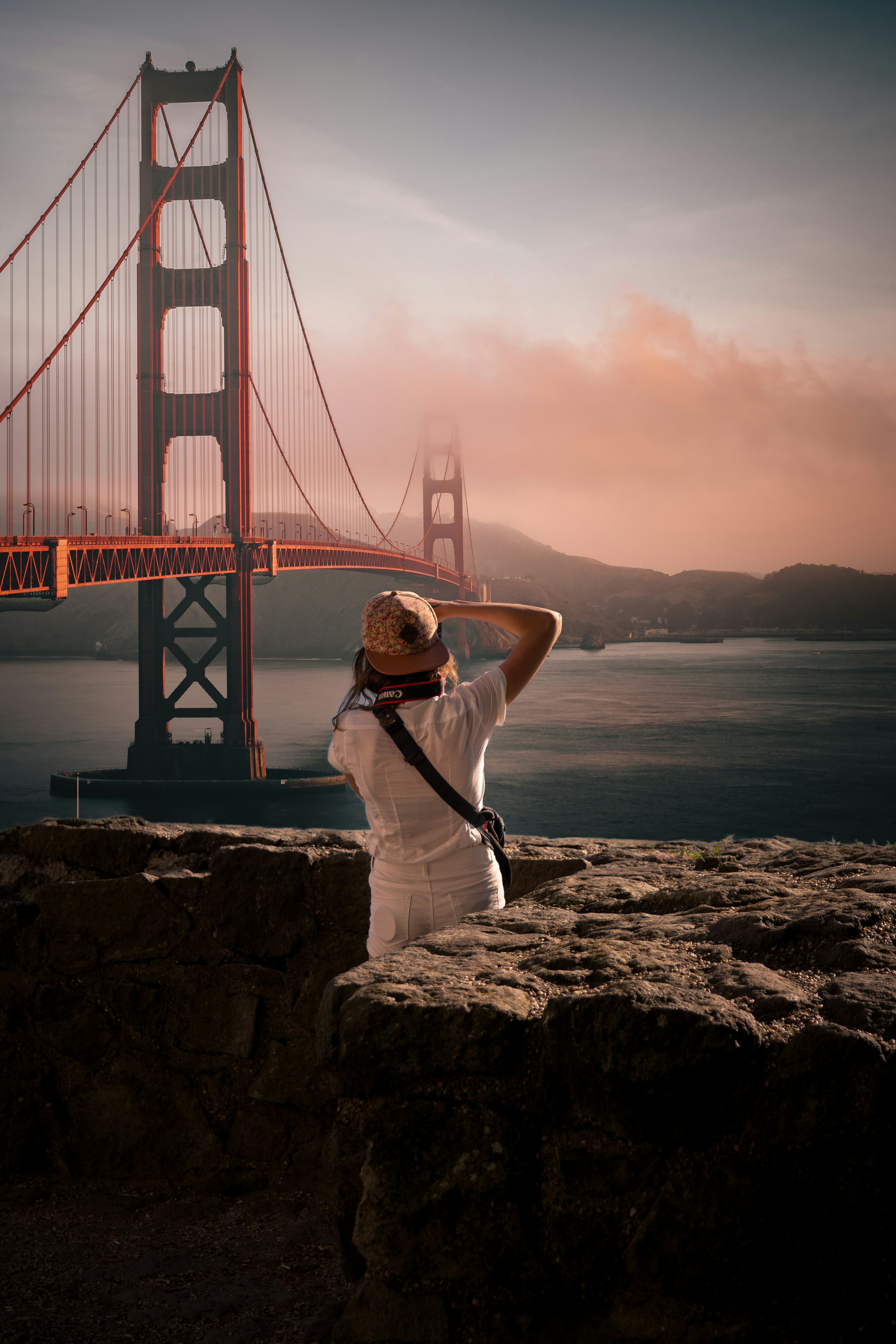 Tourist capturing the Golden Gate Bridge at dusk from a rocky viewpoint, highlighting the iconic structure against a misty backdrop.