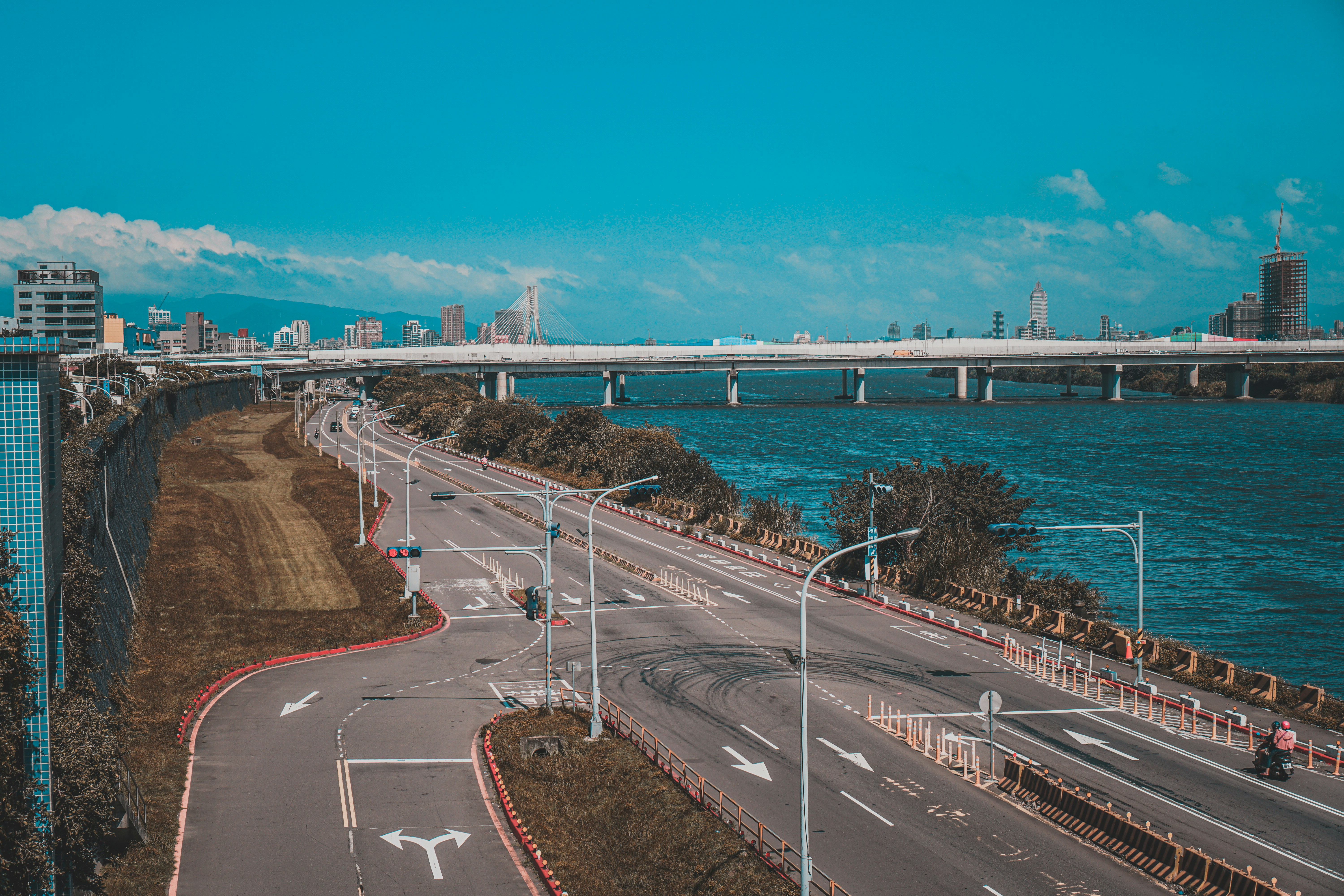 A winding road alongside a river, framed by city buildings and a bridge in the background. The scene captures the harmonious blend of urban infrastructure and natural waterway.