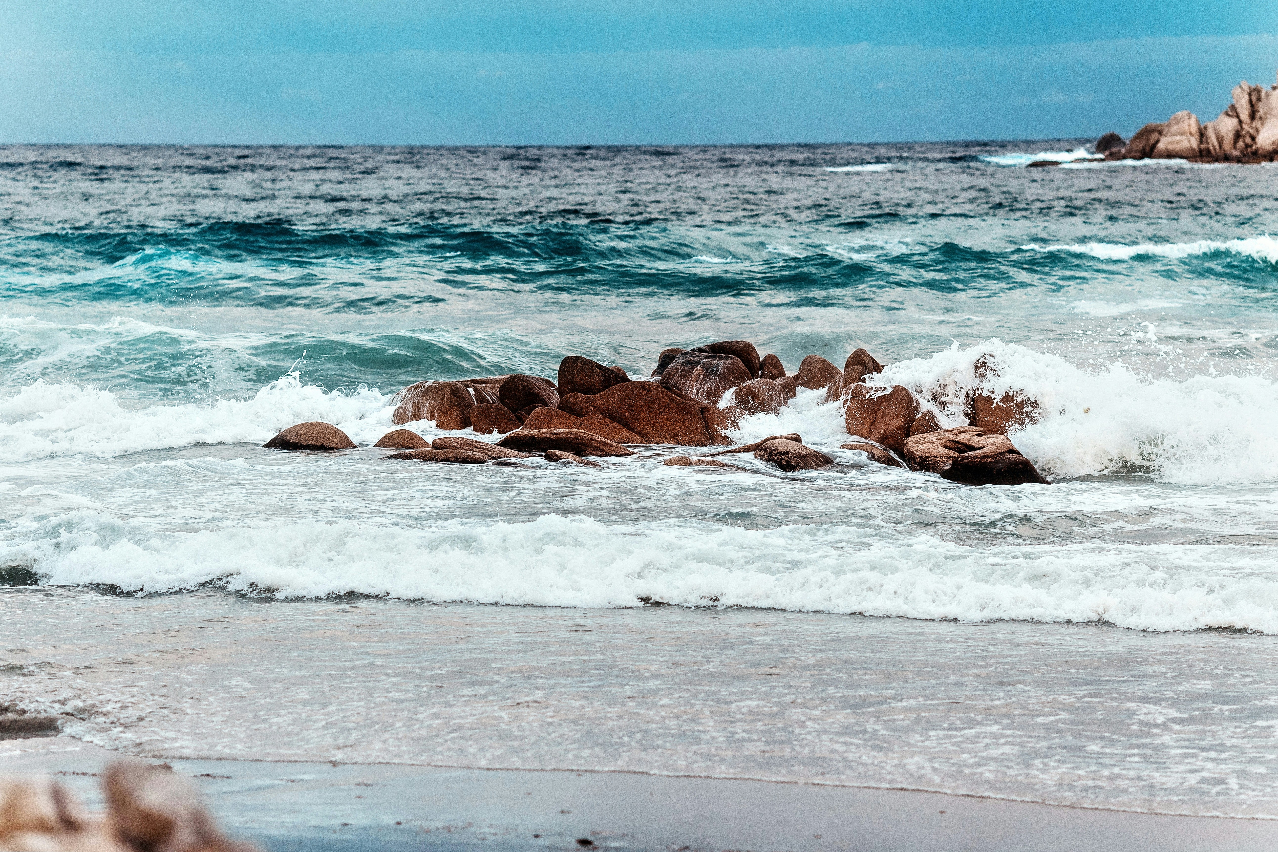 Waves gently lap against a cluster of rocks on a serene beach, under a moody sky. The interplay of water and stone creates a tranquil coastal scene.