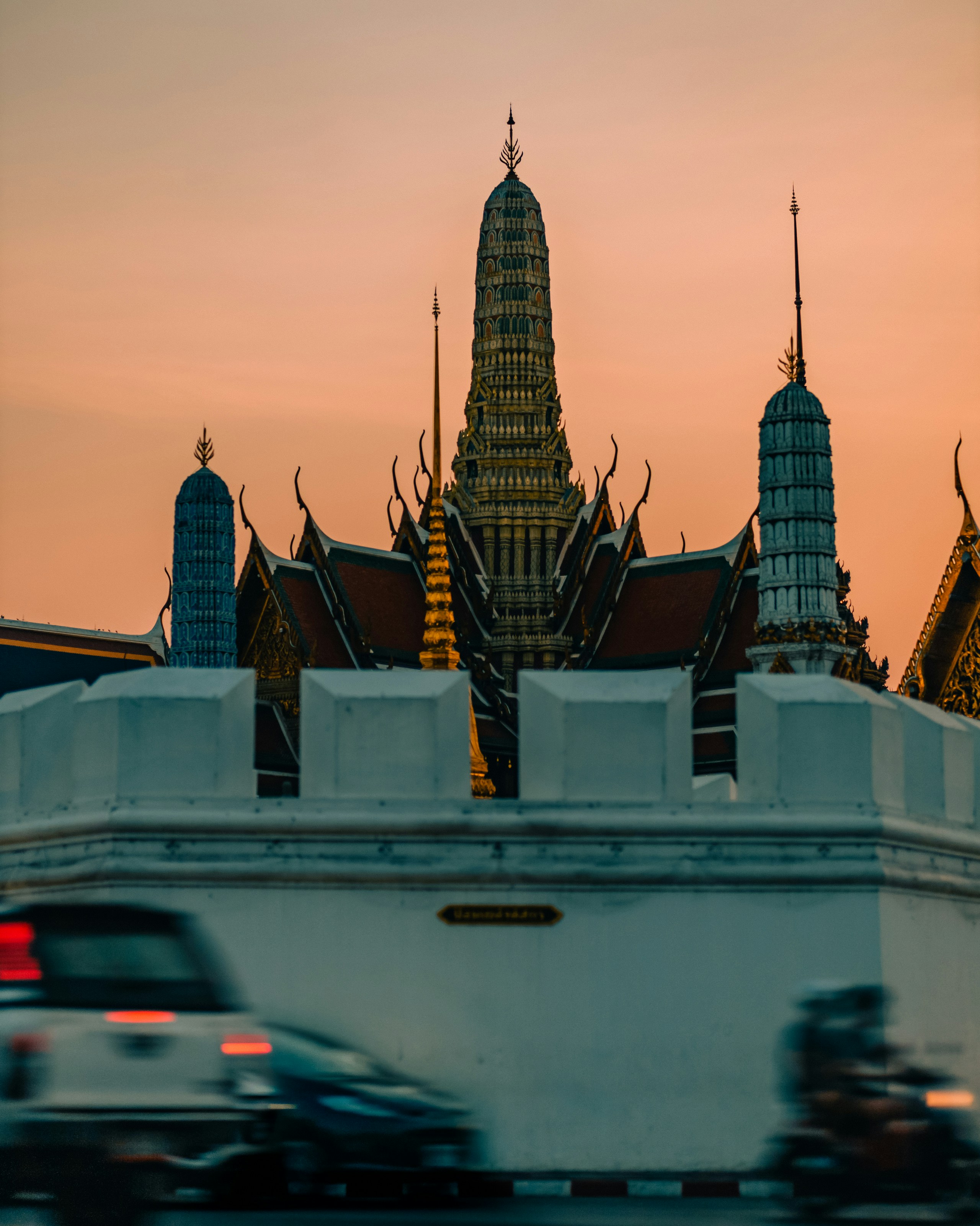 Ornate temple spires rise behind a white fortress wall under a twilight sky, with blurred vehicles passing by.