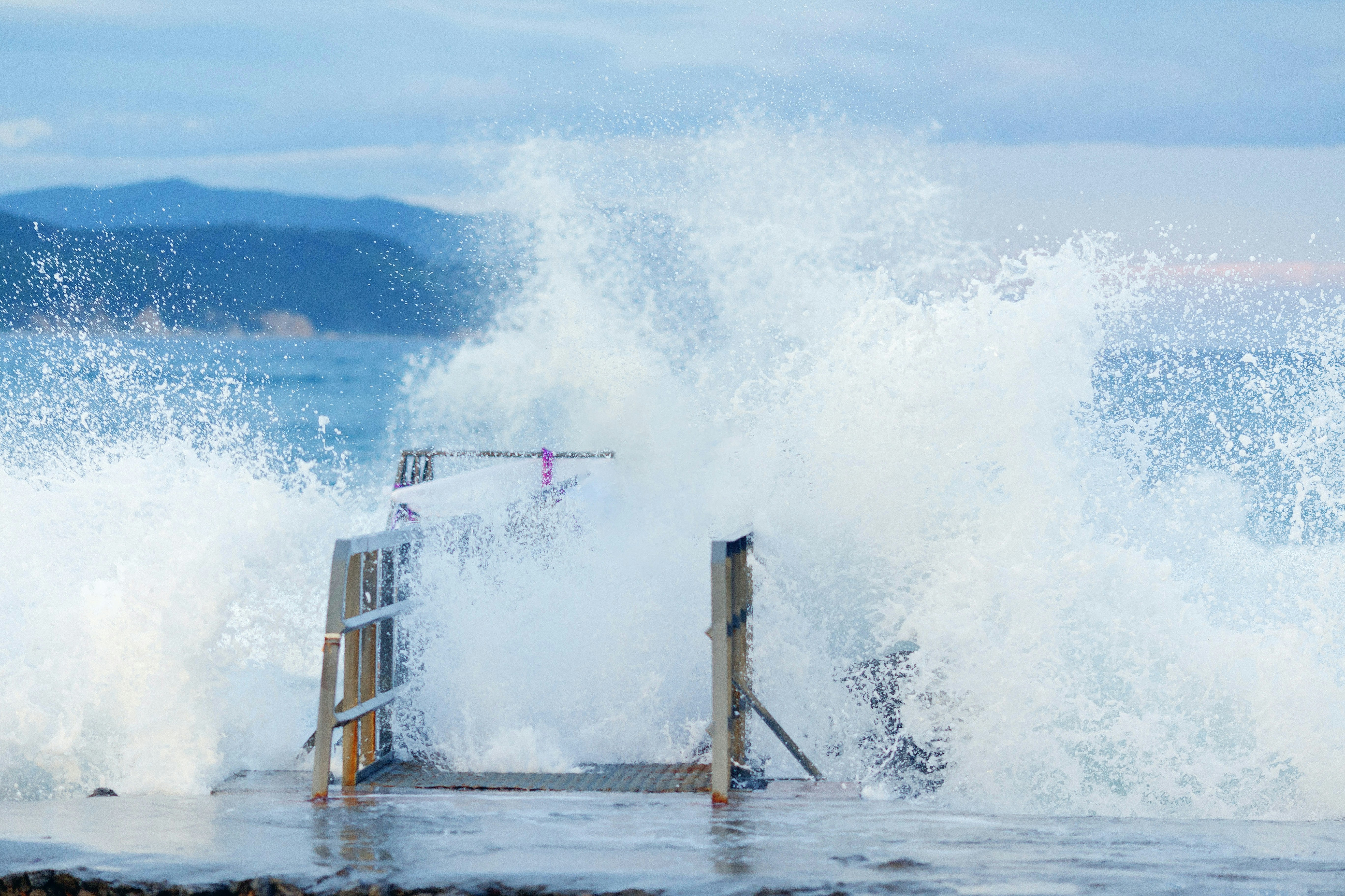 Powerful ocean waves crashing against a wooden platform, creating a spray of water against a serene backdrop of distant mountains.
