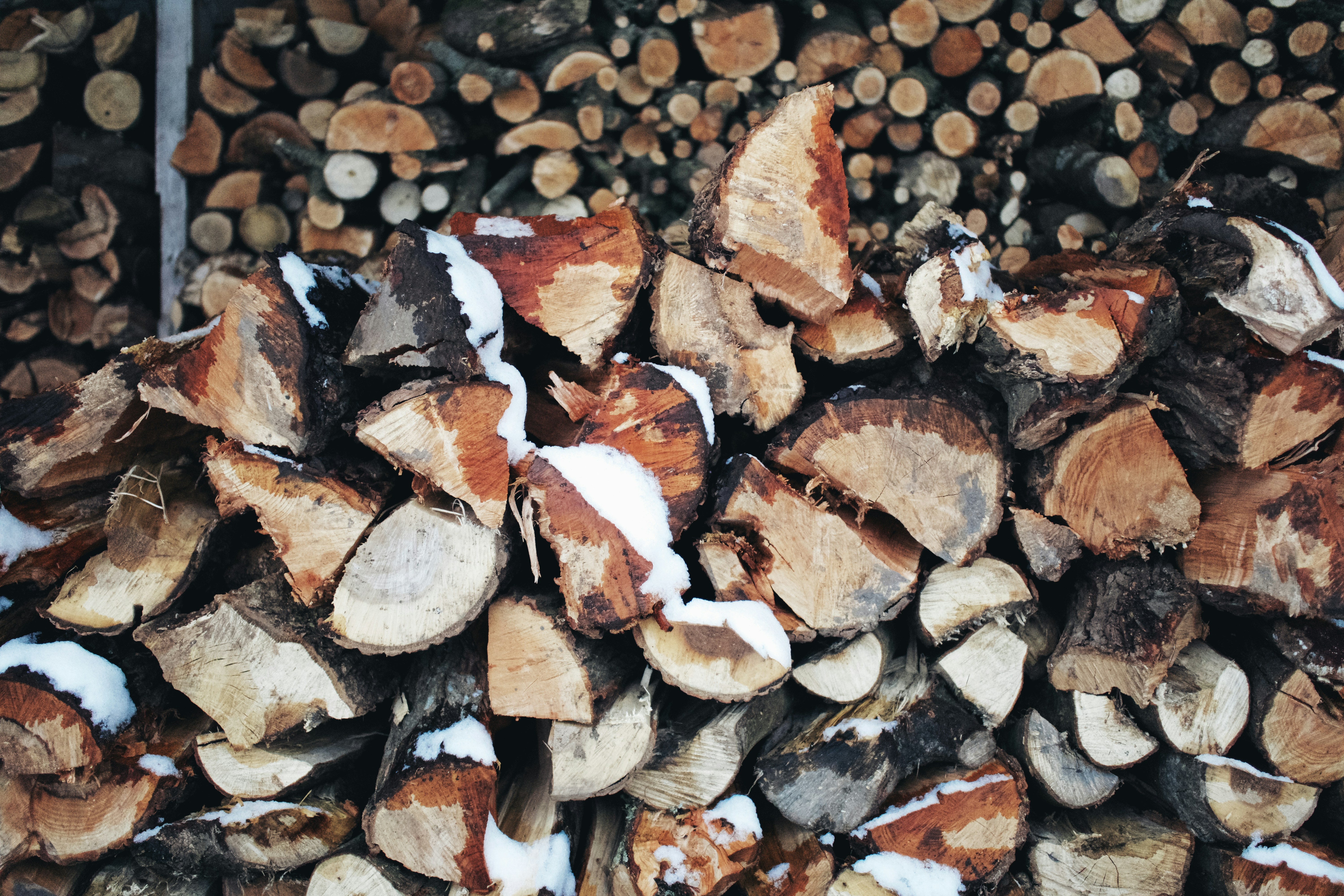 A neatly arranged pile of firewood, showcasing the textures and colors of various logs, some dusted with snow. The composition emphasizes the rustic charm of natural materials.