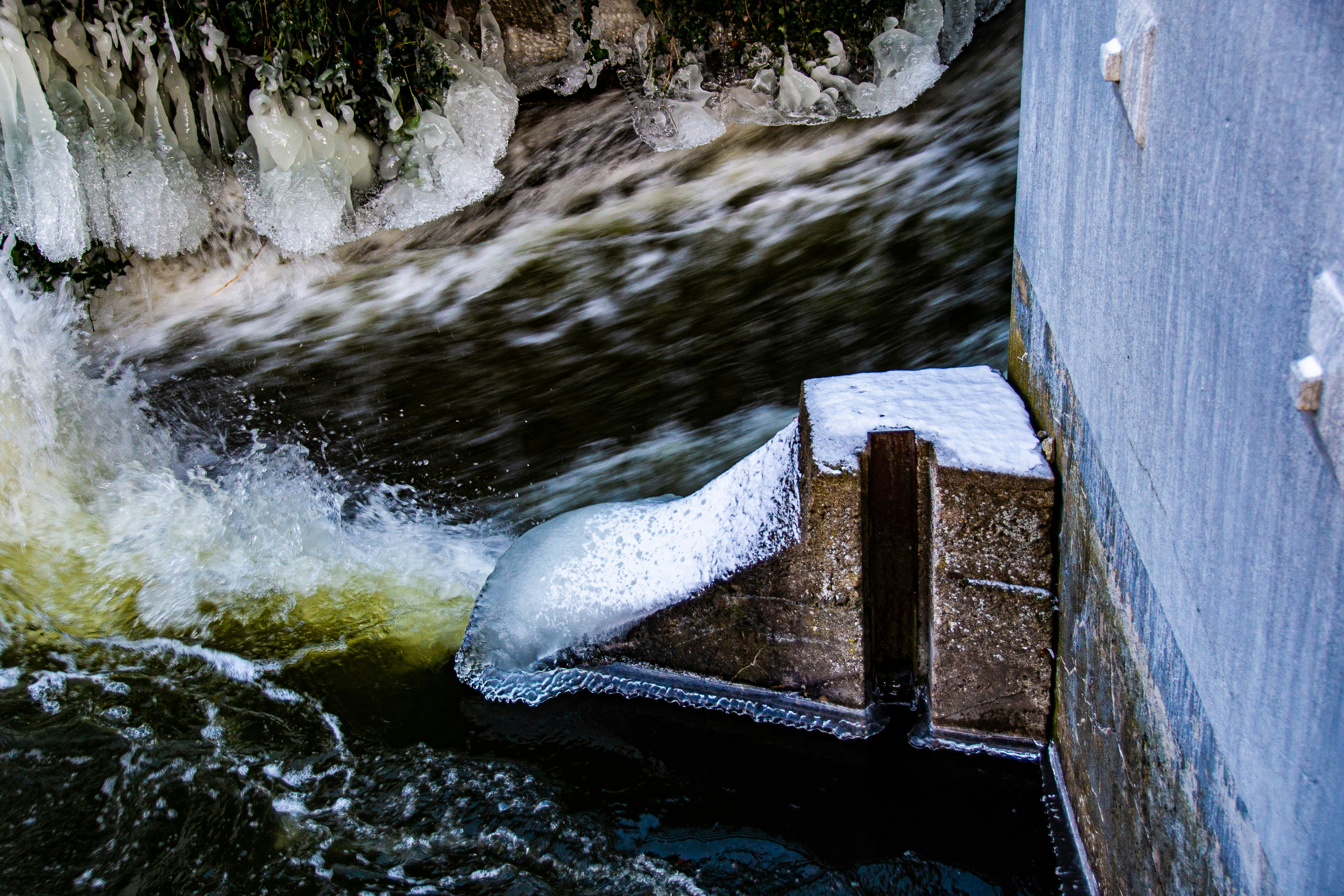 Rushing water cascades over a stone structure, revealing icy formations along the edges. The dynamic movement contrasts with the stillness of the surrounding elements.