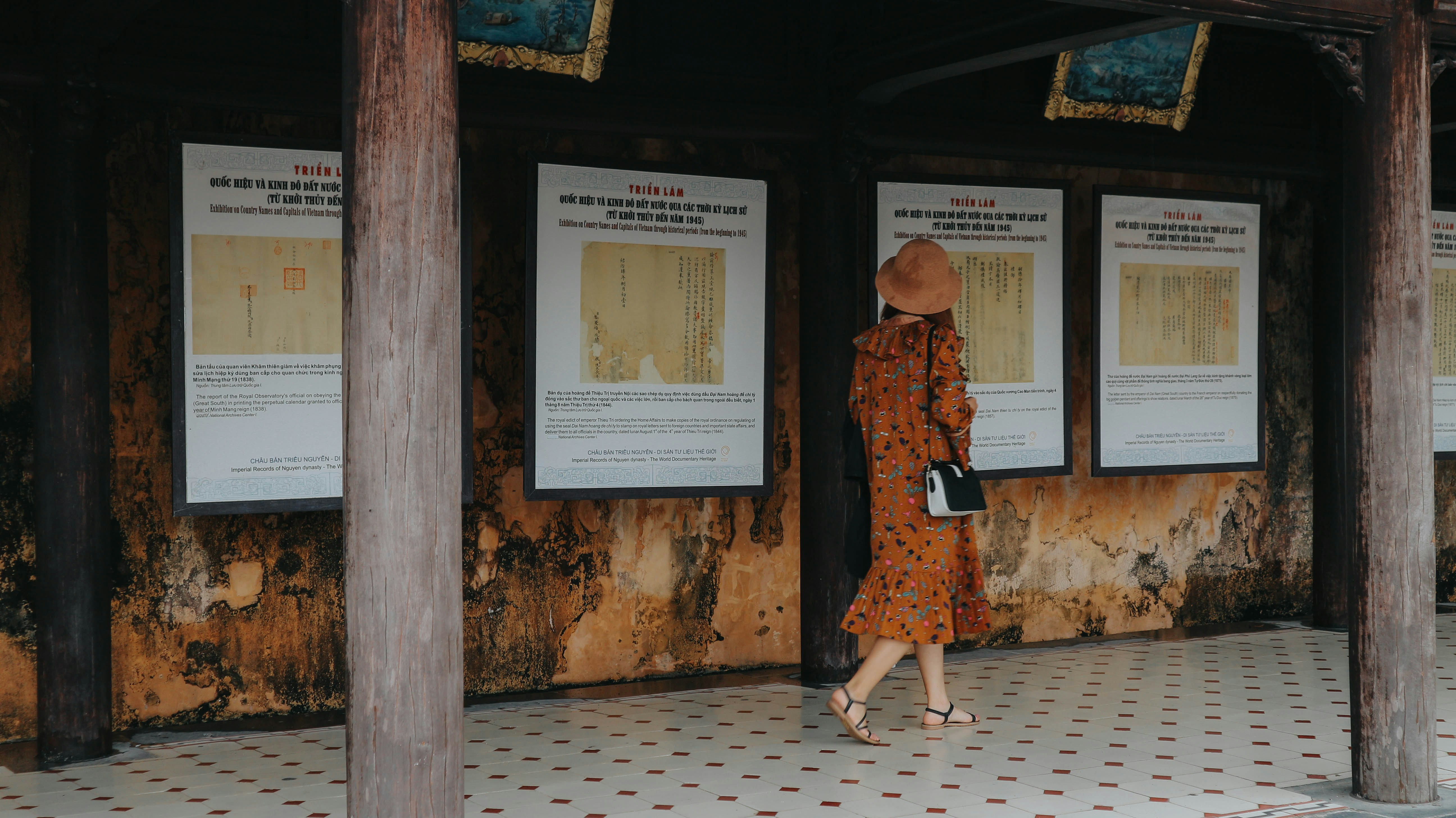 Woman in a patterned dress and hat walks past historical displays in a traditional setting. The atmosphere evokes a sense of nostalgia and exploration.