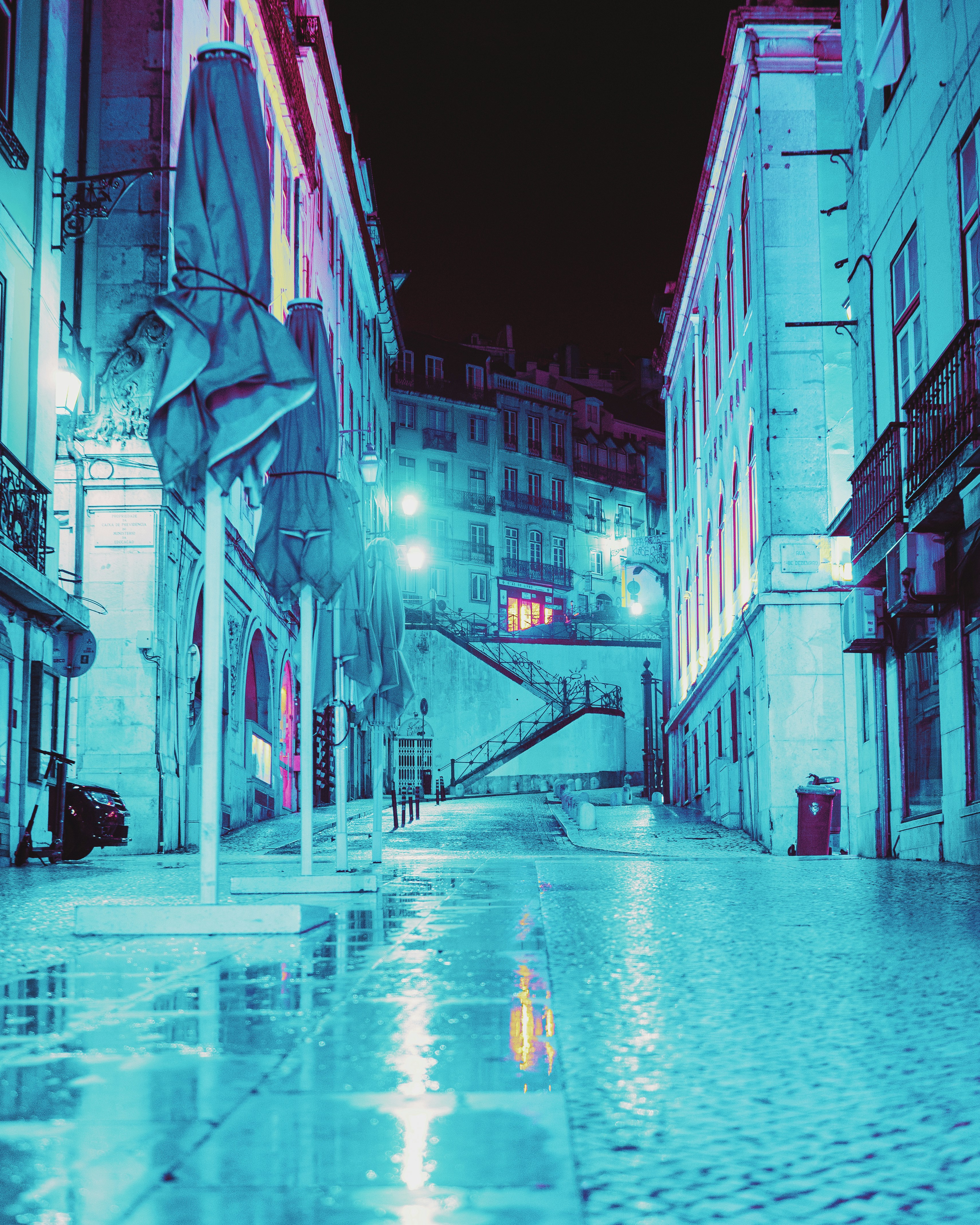 Night street scene bathed in neon blue light, with wet cobblestones reflecting street lamps and a distant stairway between buildings.