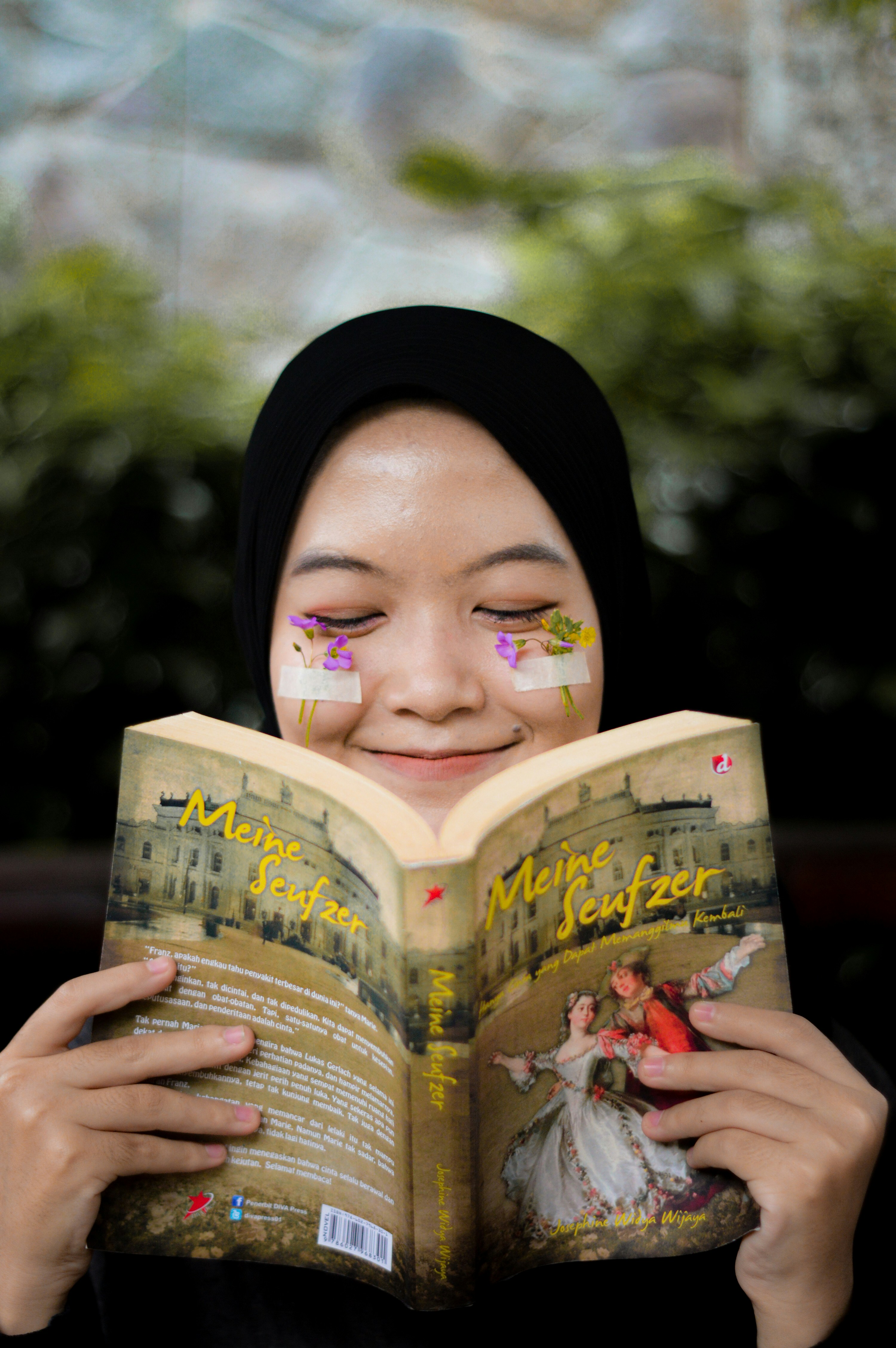 Young woman reading 'Meine Seyfer' with floral-themed face art, surrounded by a lush green background.