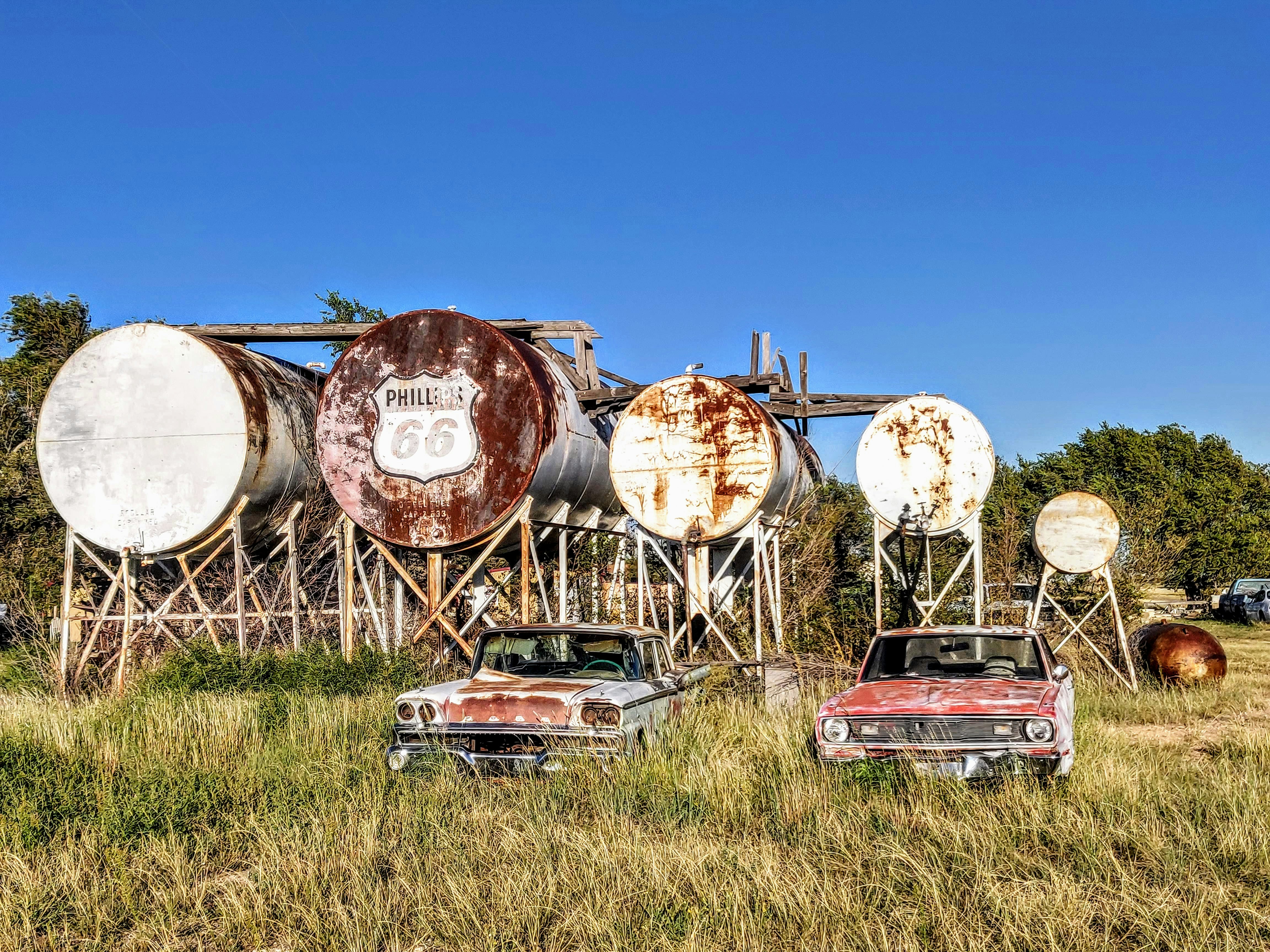 Two red and white vintage cars parked in front of weathered silos on a grassy field under a clear blue sky.