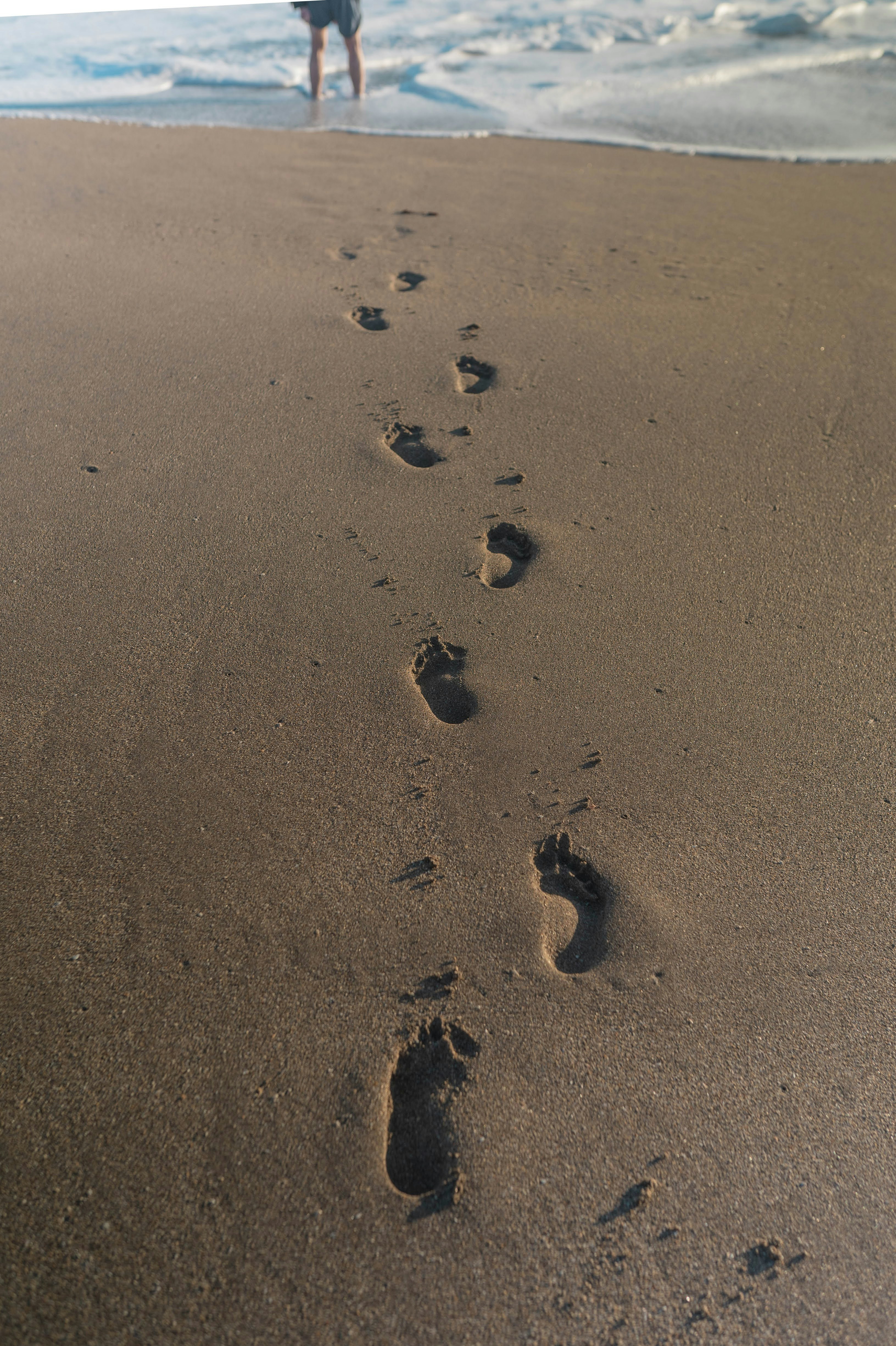 Footprints leading toward the ocean on a sandy beach, with gentle waves lapping at the shore in the background.