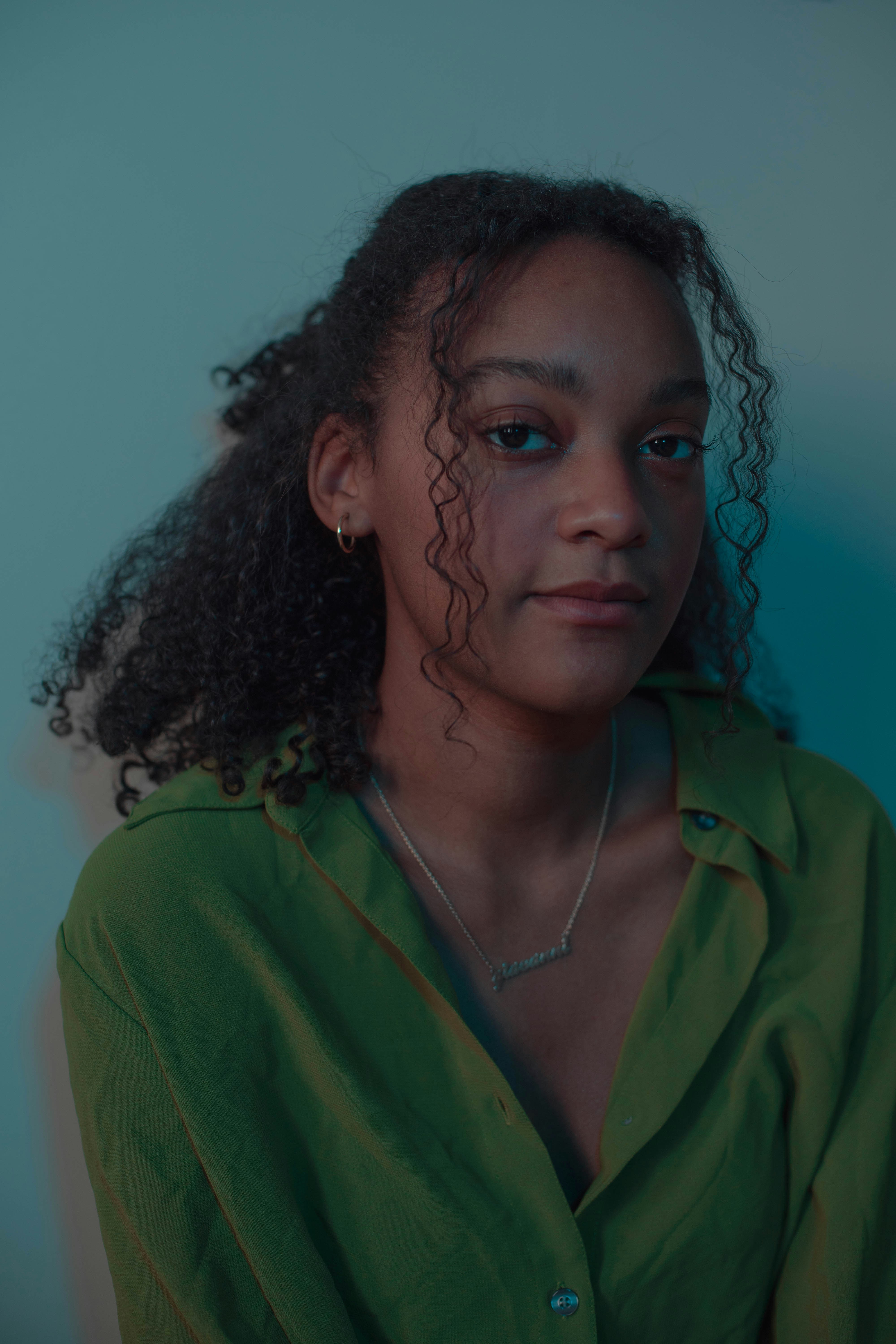 Portrait of a young woman with curly hair, wearing a green shirt, against a softly lit backdrop. The subtle play of colors enhances her serene expression.