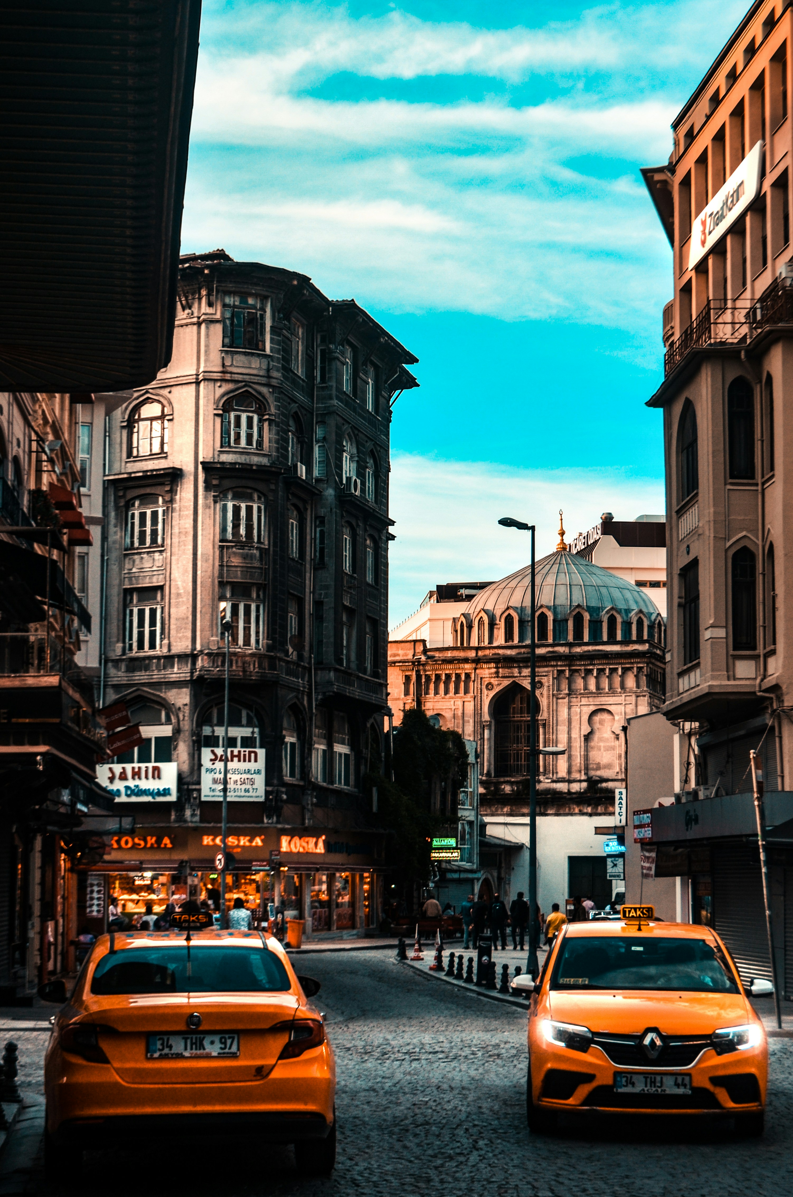 A vibrant street scene featuring yellow taxis navigating a cobblestone road, framed by historic buildings and a domed structure in the background.