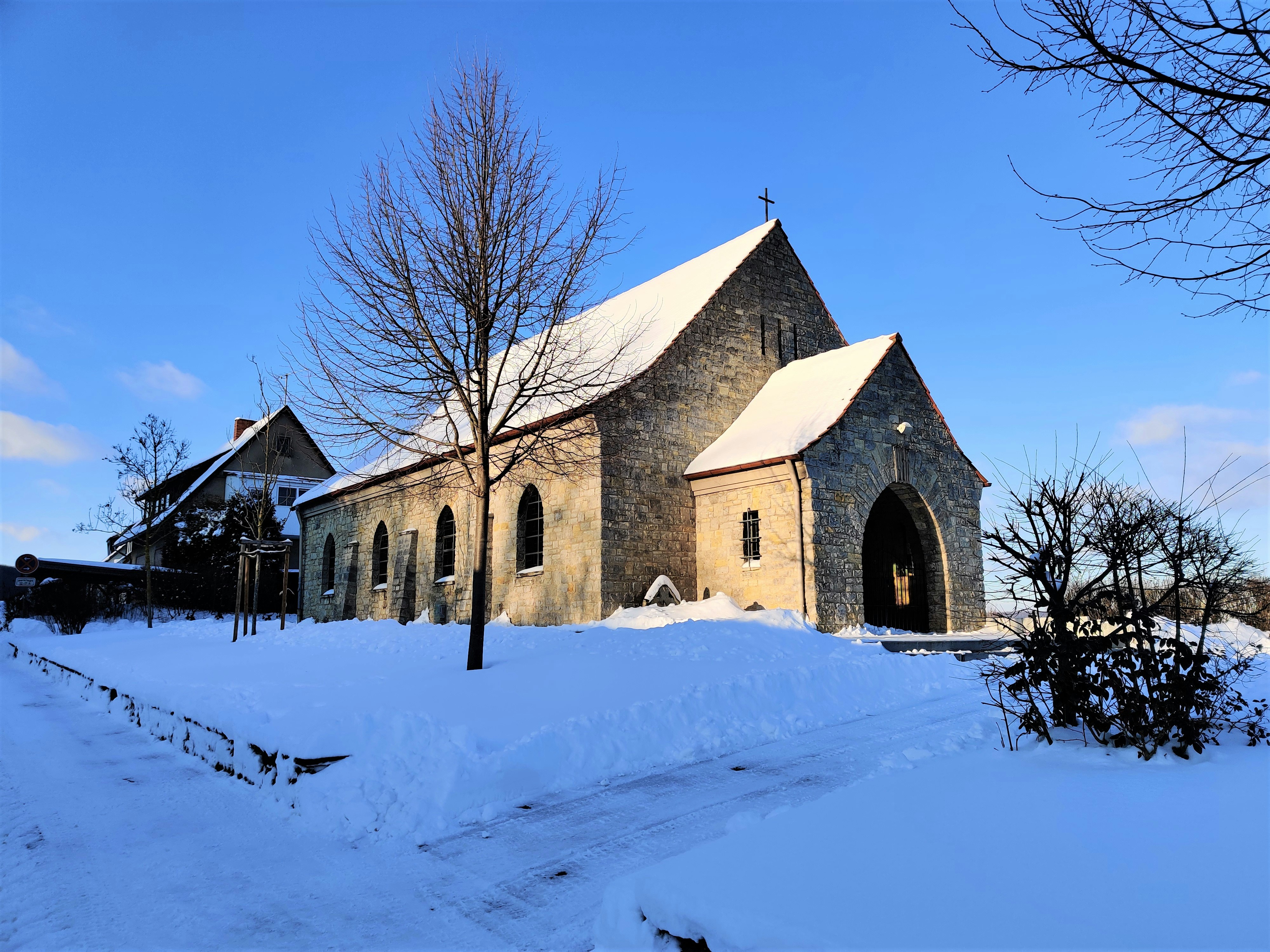 Snow-covered stone church under a bright blue sky with bare trees. A snow-filled path leads to the arched entrance.