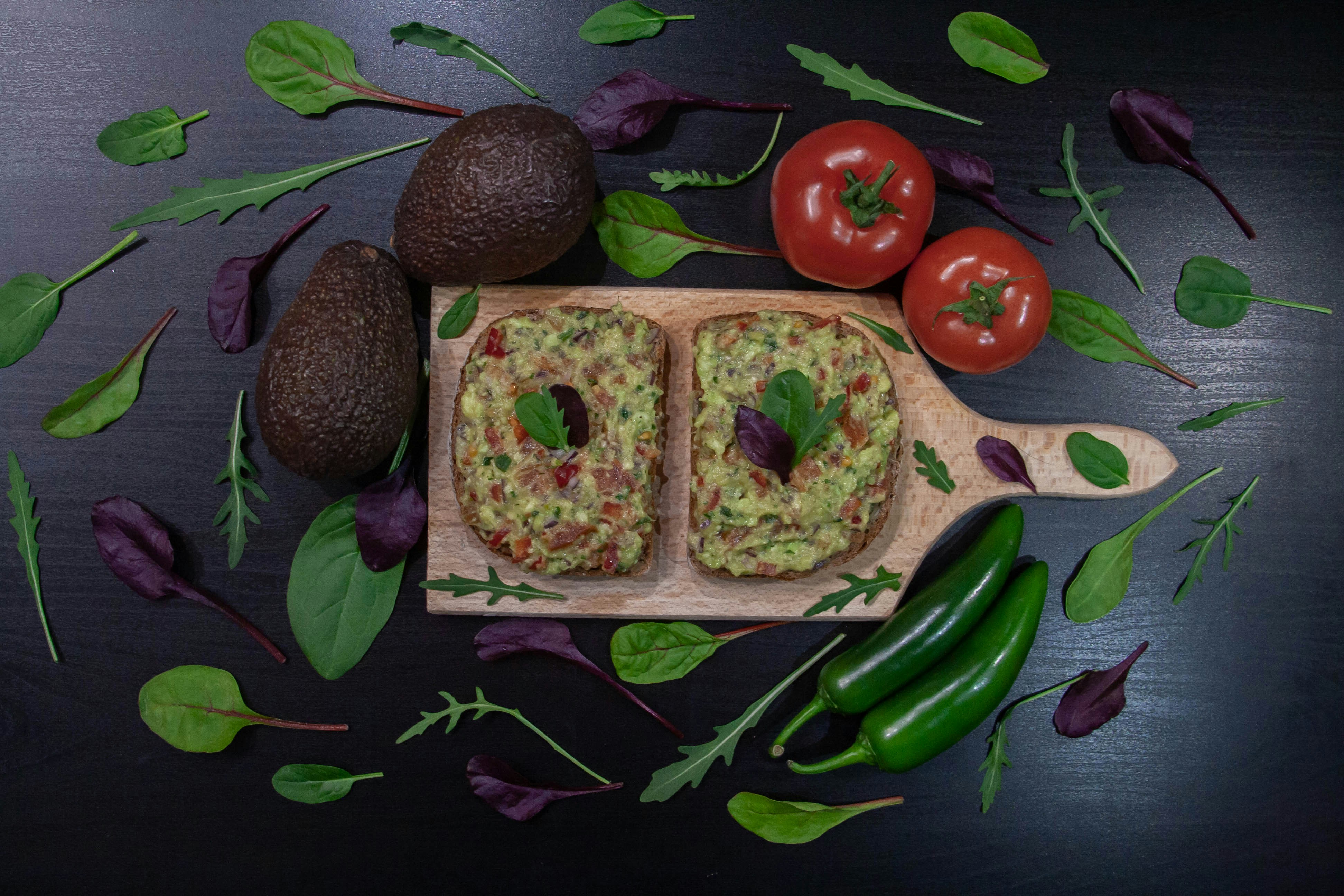 Avocado toast on a wooden board surrounded by fresh tomatoes, jalapeños, and leafy greens.