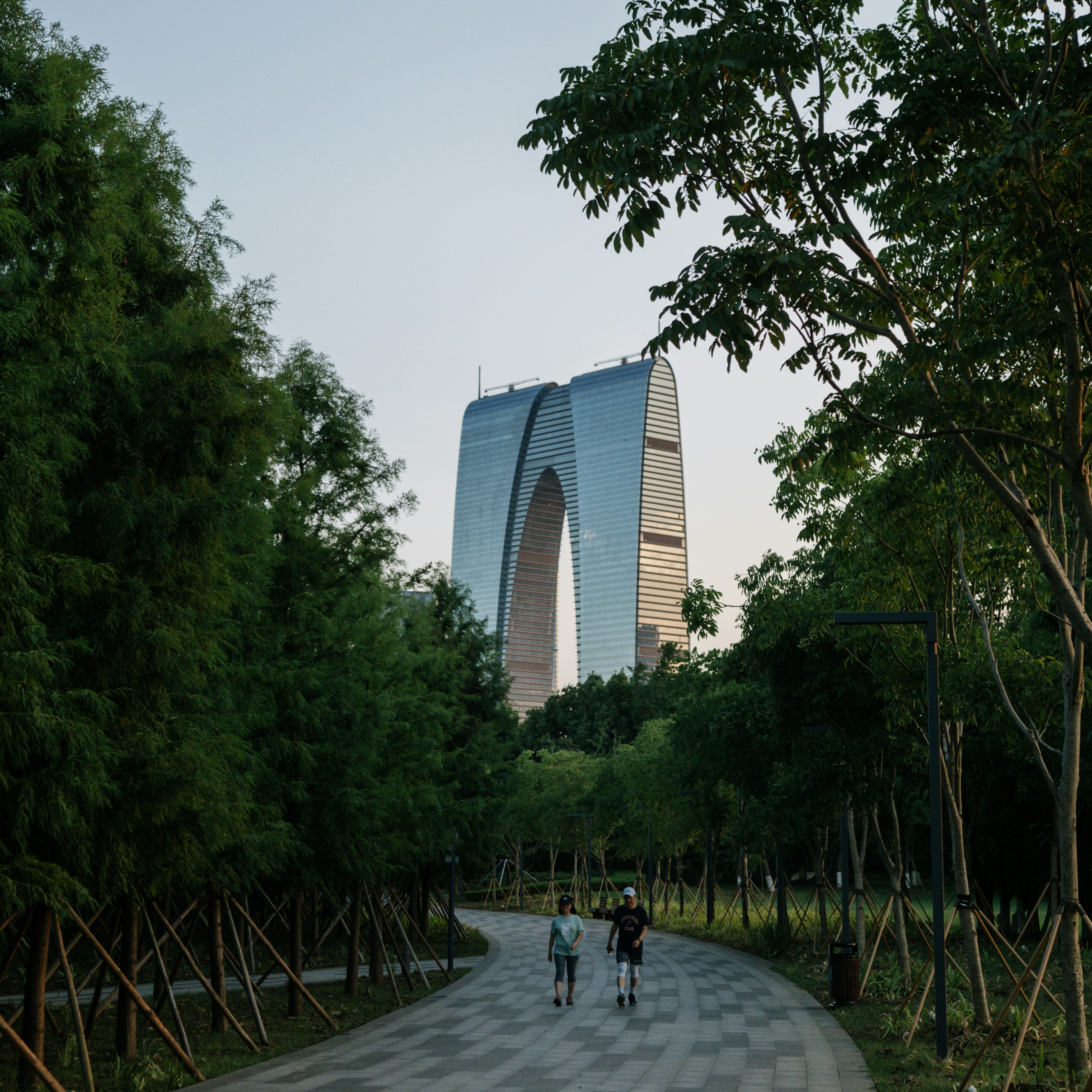 A photograph capturing a tree-lined park path with a futuristic glass-arch tower rising in the distance.