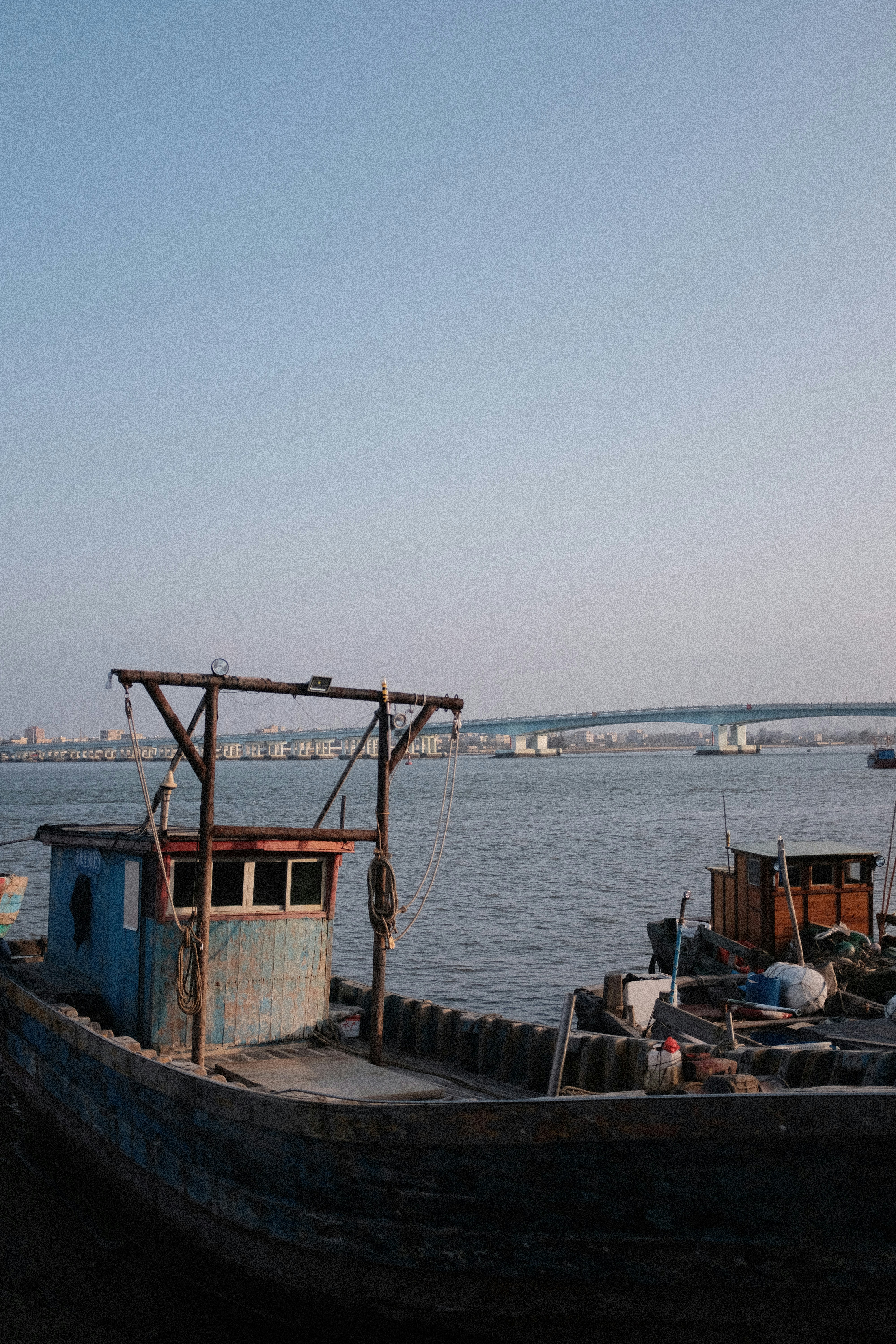 Weathered fishing boat docked by the river, surrounded by remnants of daily life and a distant bridge. 