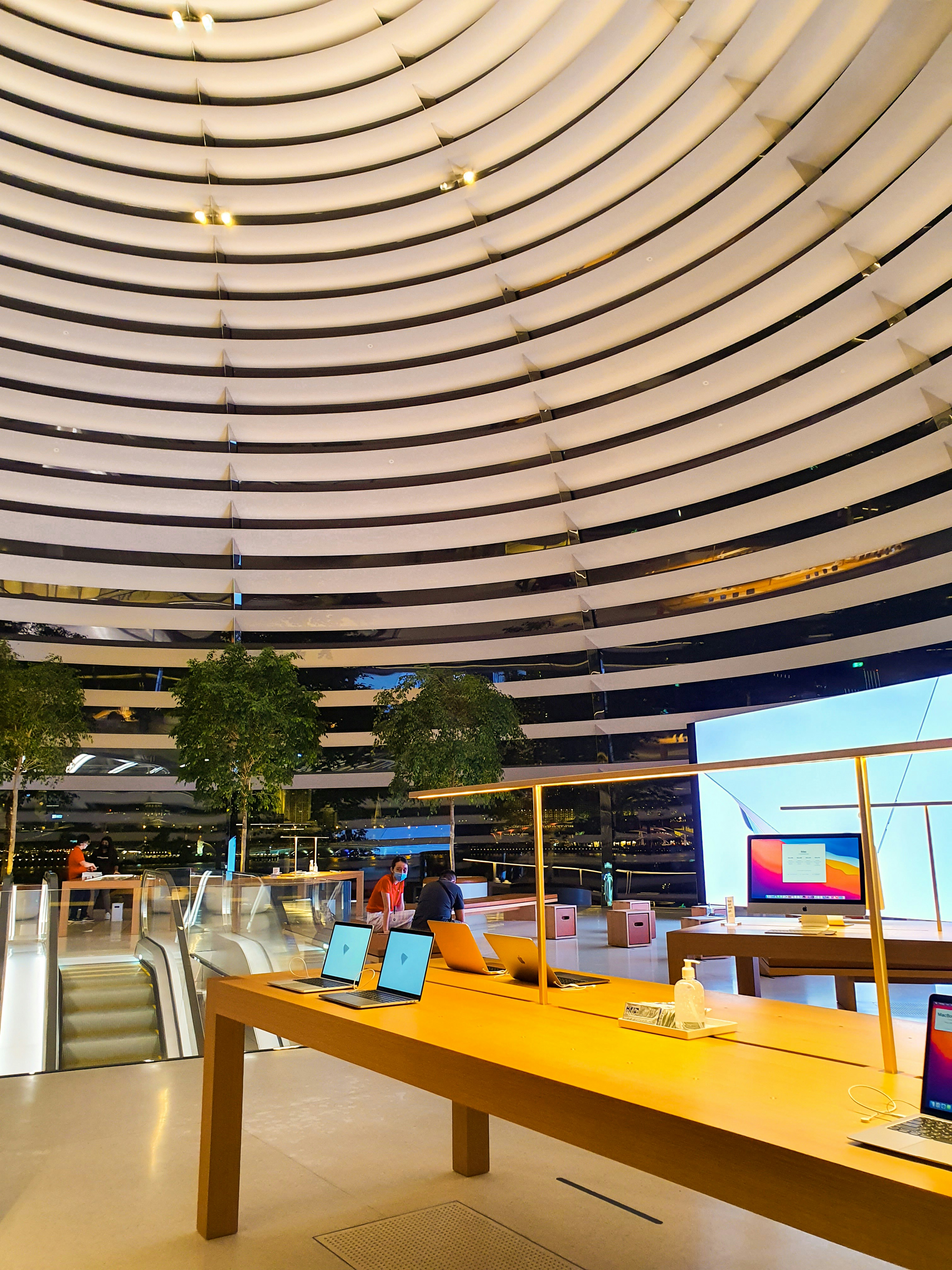 Laptops line a long wooden table inside a circular tech showroom beneath a concentric, ringed ceiling. Greenery and display screens punctuate the modern, open space.