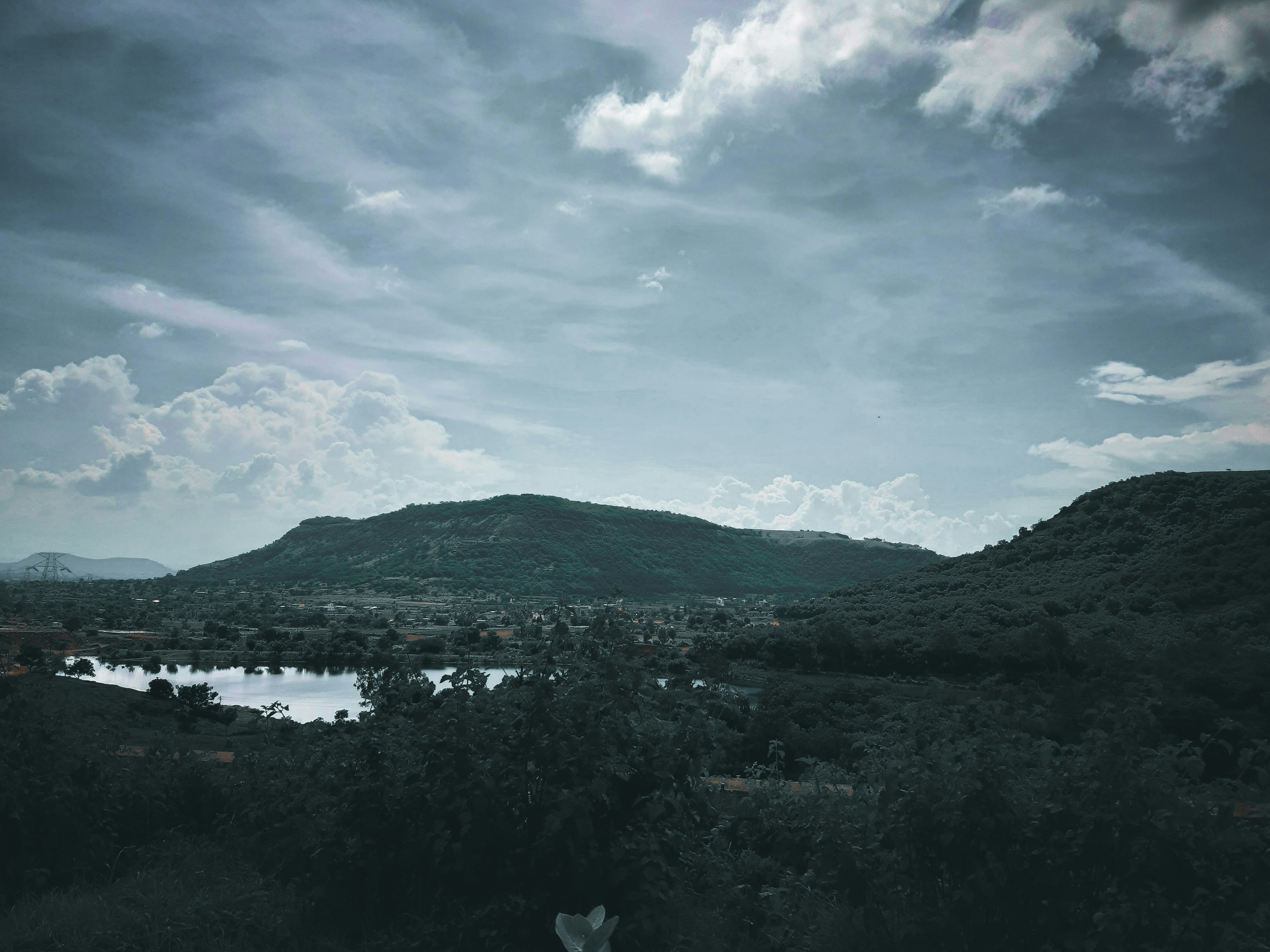 Landscape photograph of rolling hills behind a calm lake with a distant town, under a blue, cloud-filled sky.