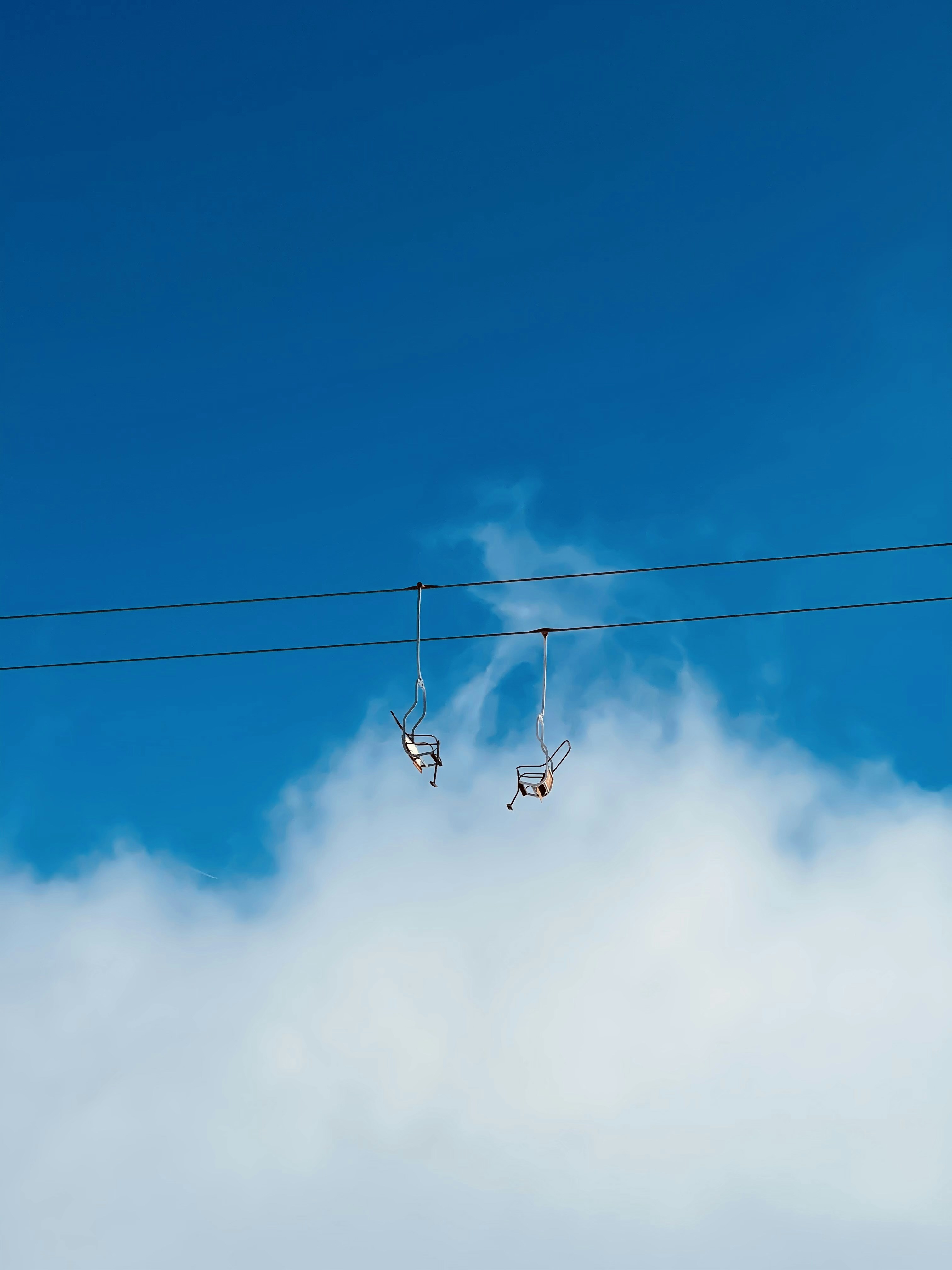 Two empty chairlifts suspended against a clear blue sky, with wisps of clouds below. A quiet moment captured in the tranquility of nature.