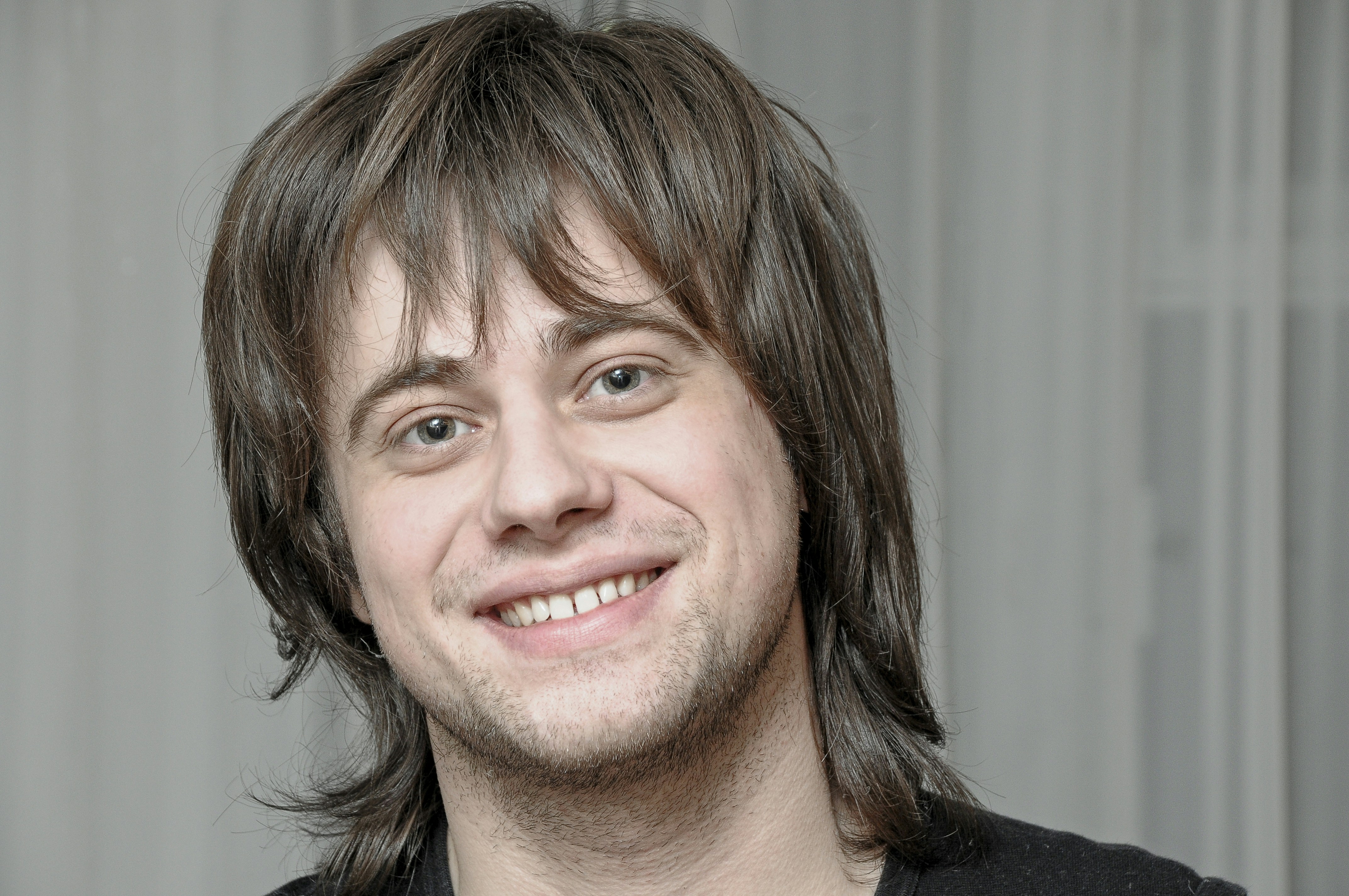 A young man with shoulder-length hair smiles warmly, showcasing a friendly demeanor against a softly blurred background.