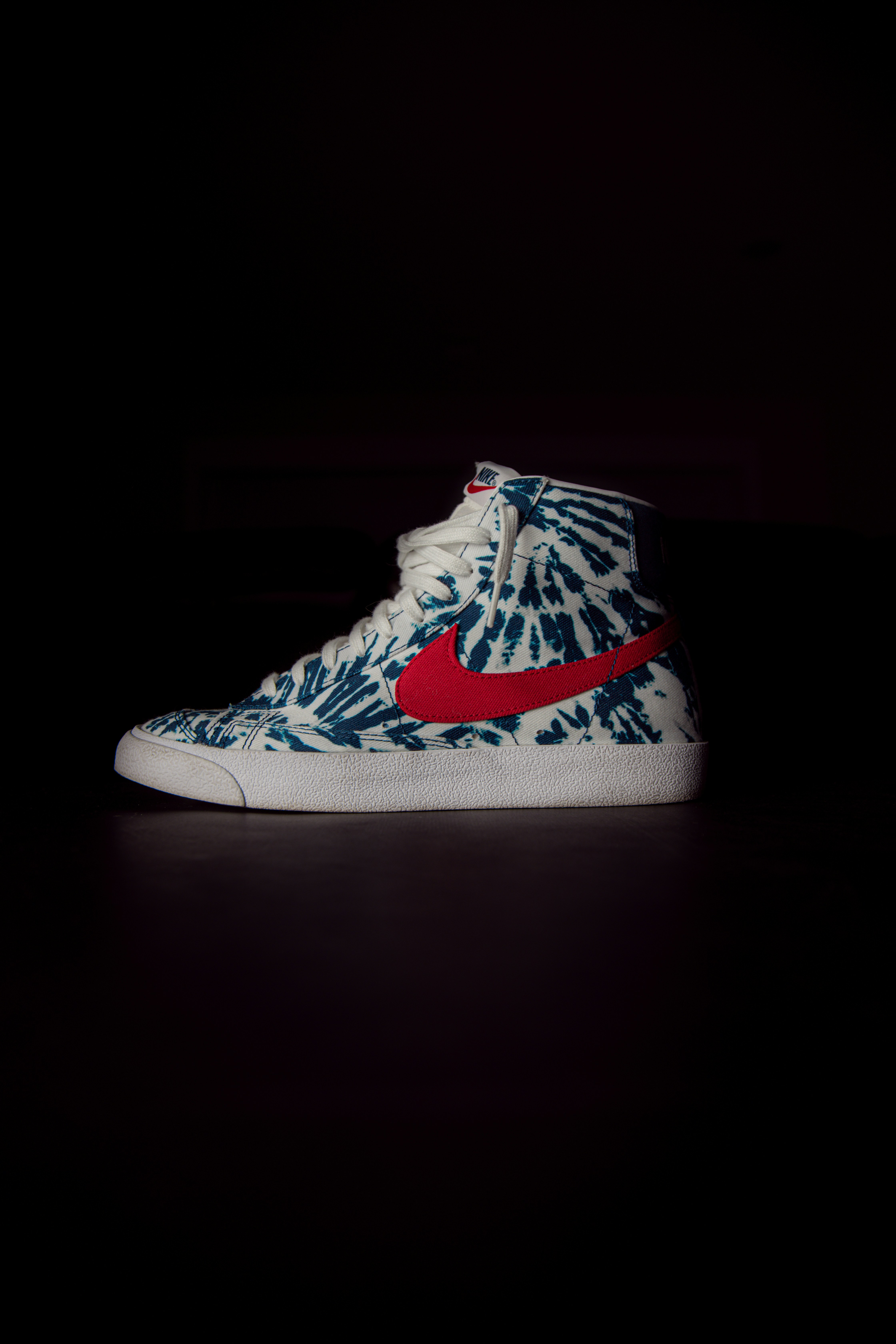 High-top sneaker featuring a unique blue and white tie-dye pattern with a bold red Nike swoosh. The shoe is set against a dark background, emphasizing its design.