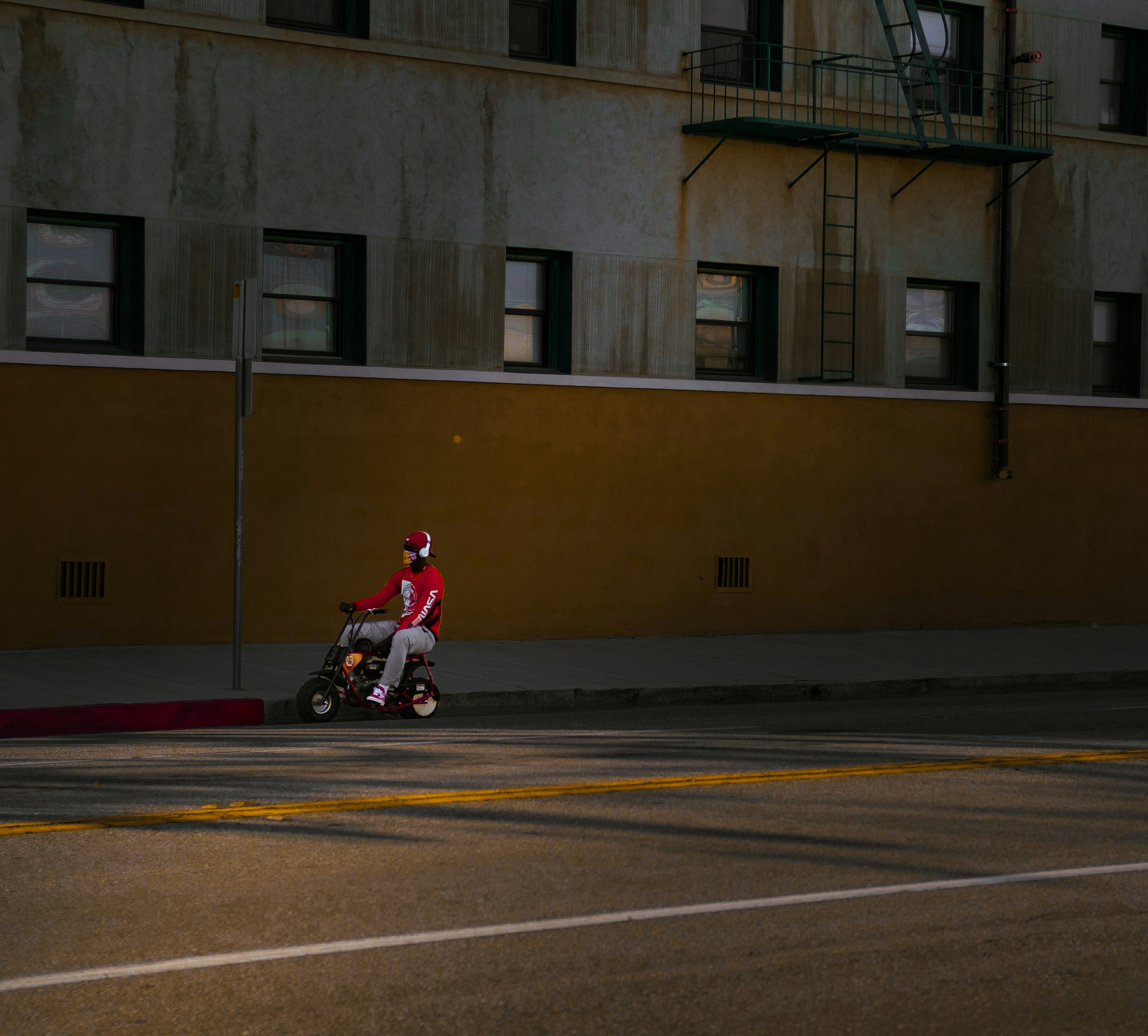 Motorcyclist in a red jacket rides along a city street beside a tall building during daylight.