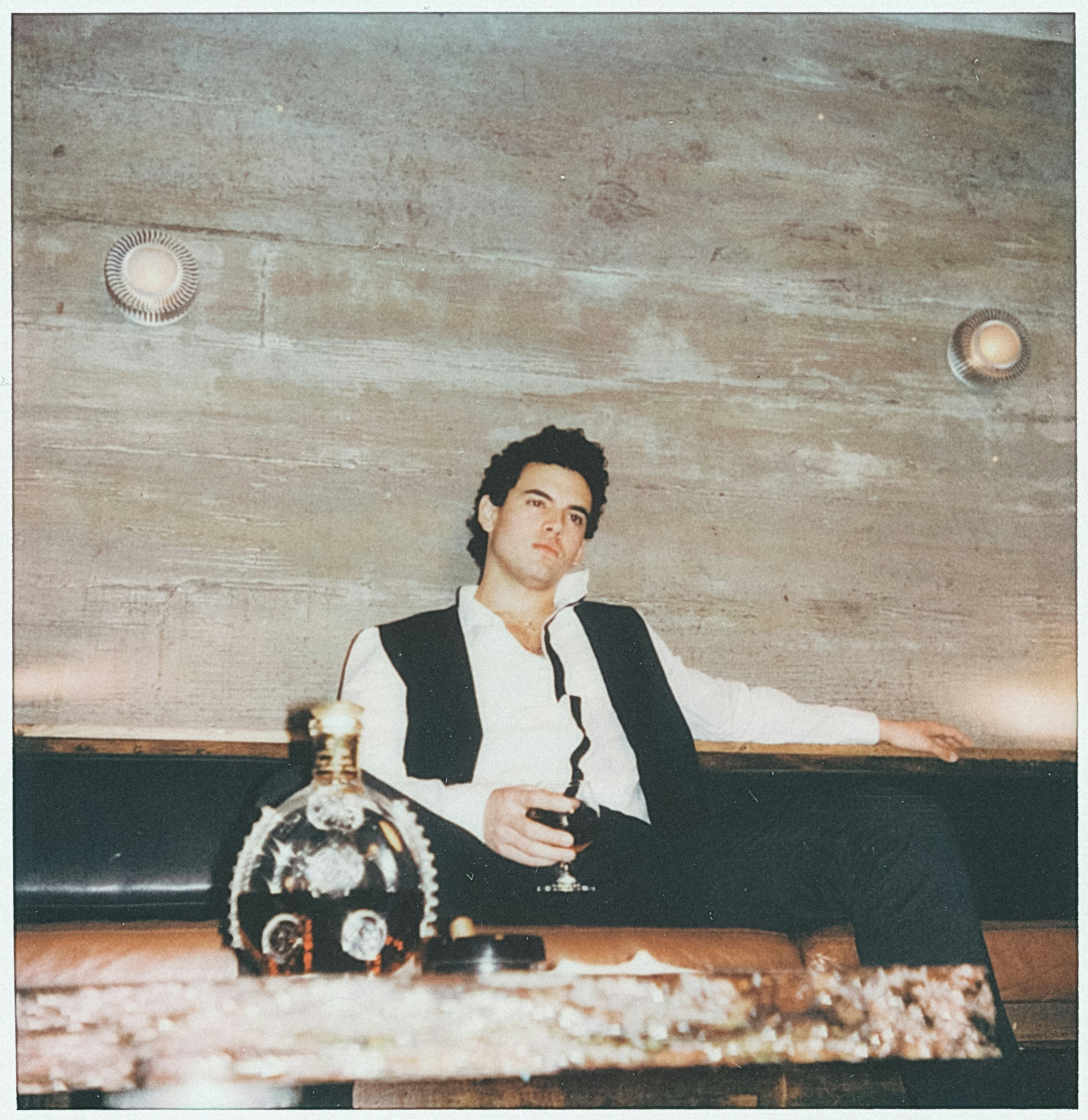 Man in a stylish outfit sitting on a leather couch, holding a glass of drink, with a bottle of cognac in the foreground. The ambiance reflects a contemporary lounge setting.
